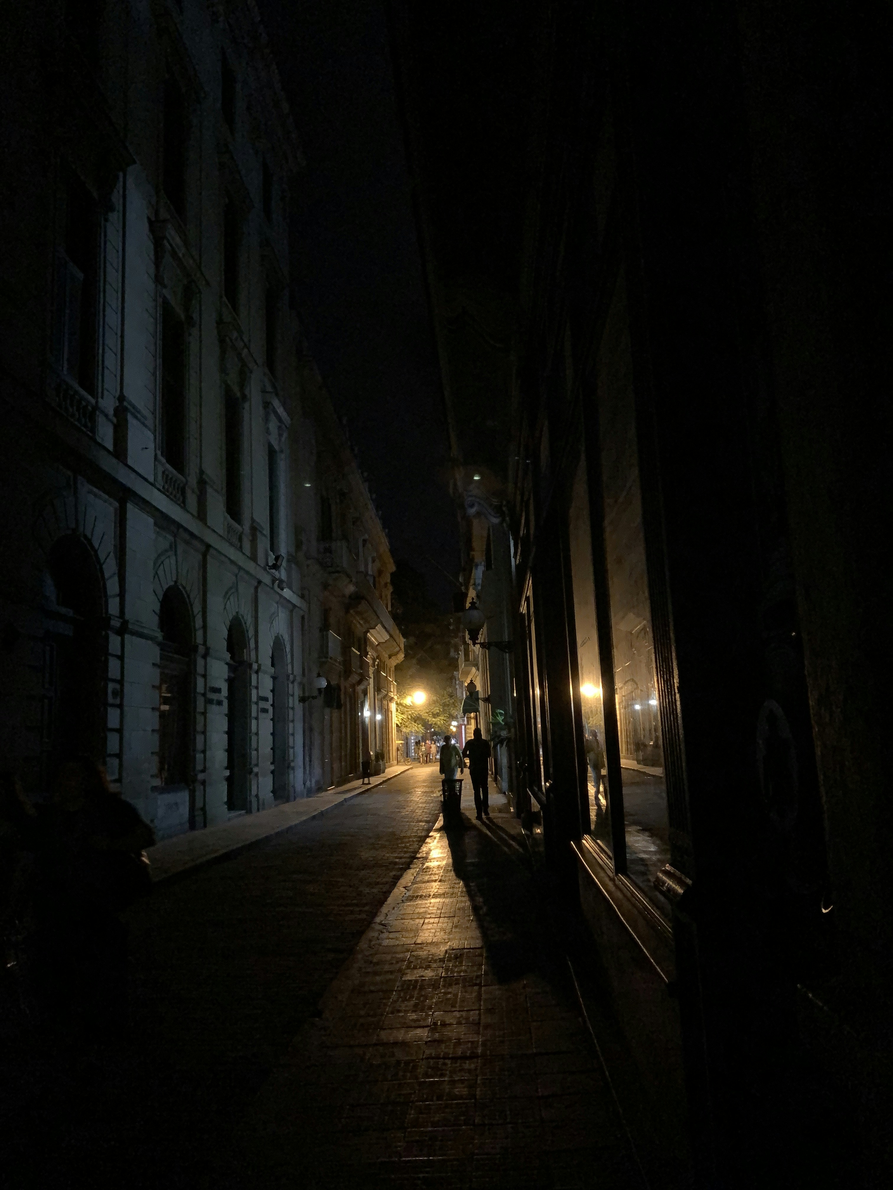 Dimly lit alleyway with historical architecture, featuring shadows and warm light from street lamps. A solitary figure stands in the distance.