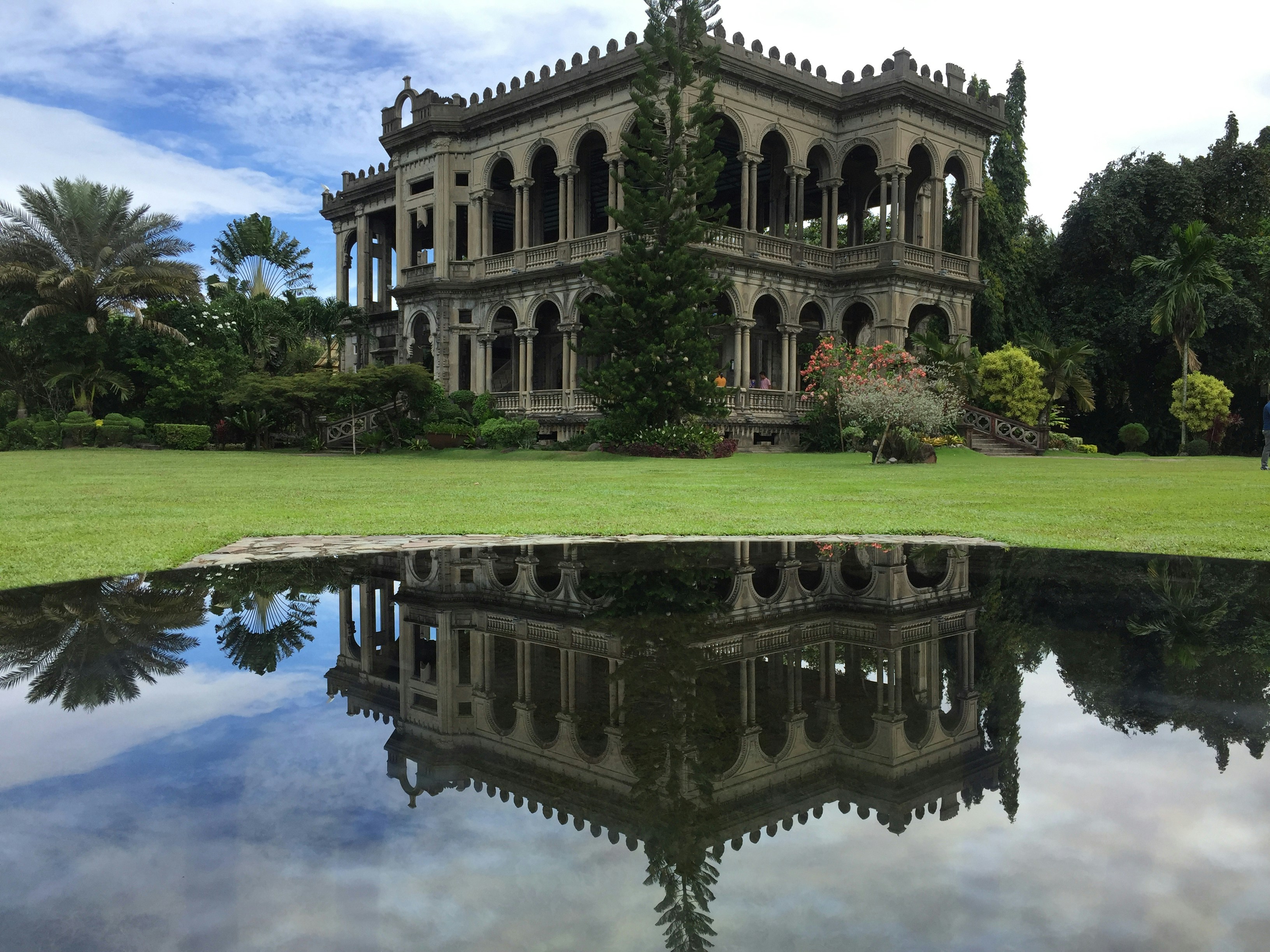 Old stone mansion reflected in a tranquil pond surrounded by lush greenery and a partly cloudy sky.