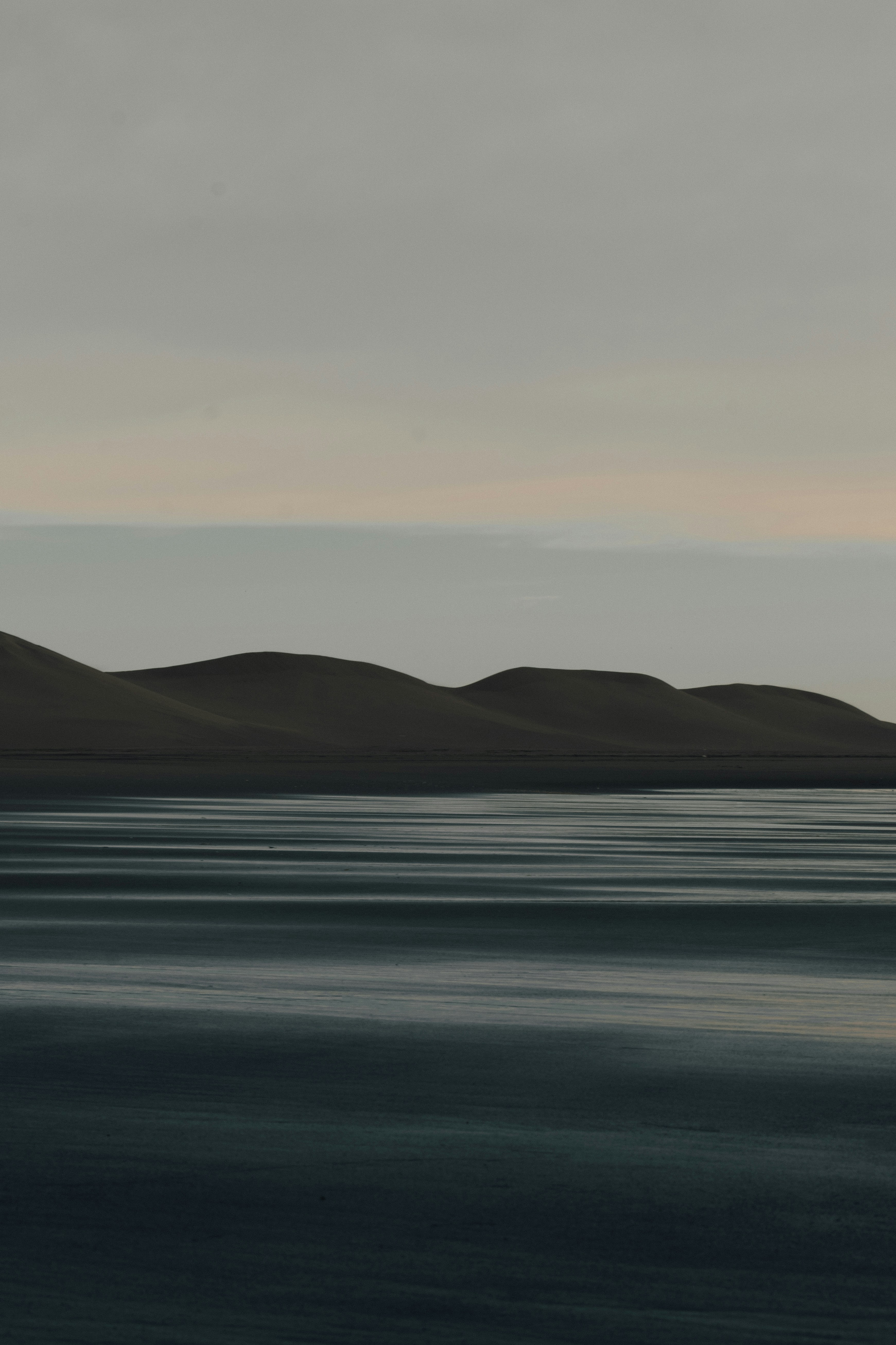 Gentle waves ripple across a serene body of water, reflecting the subtle contours of distant sand dunes under a muted sky.