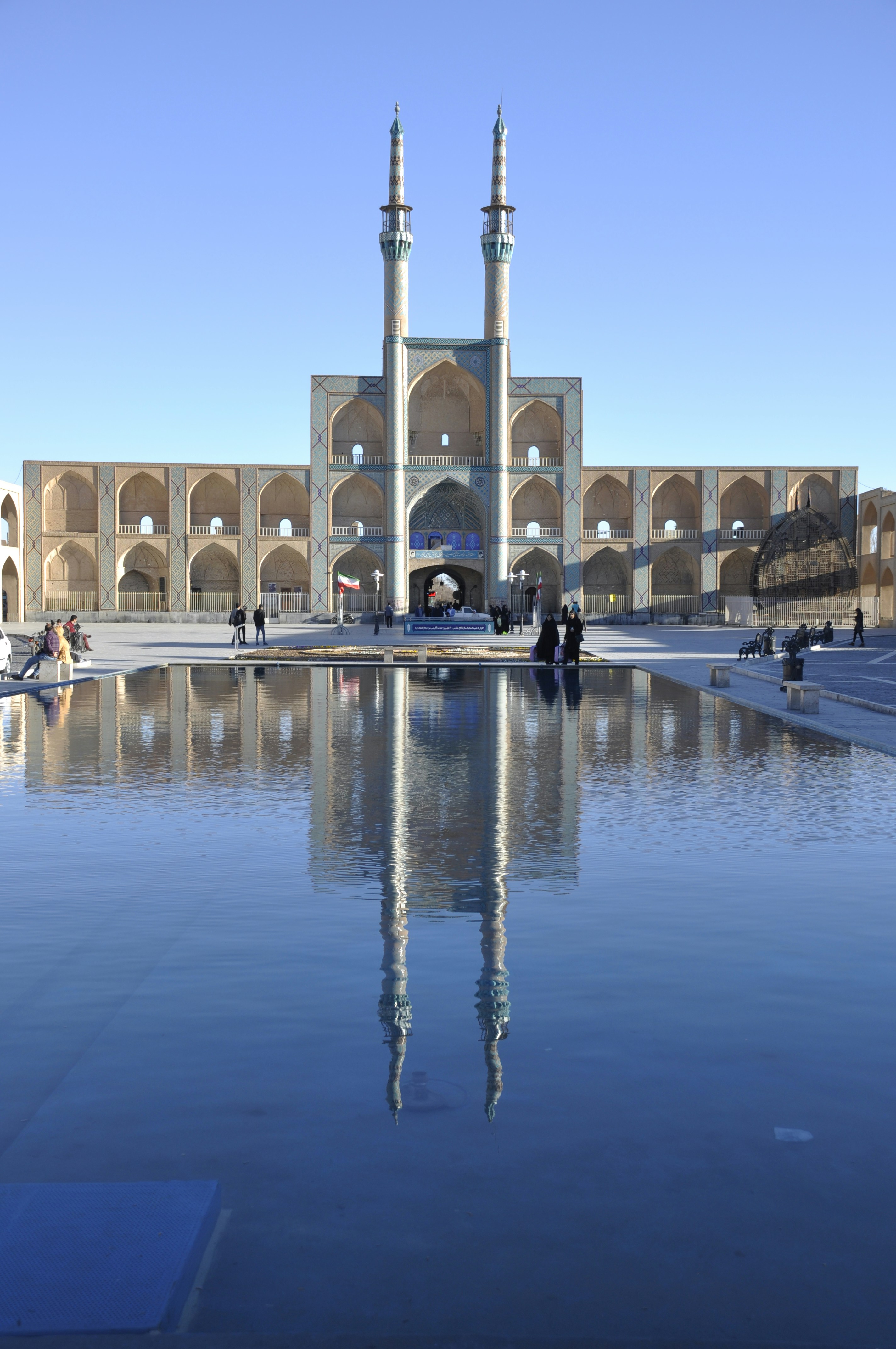 Historic mosque with twin minarets reflected in a calm pool under a clear blue sky.