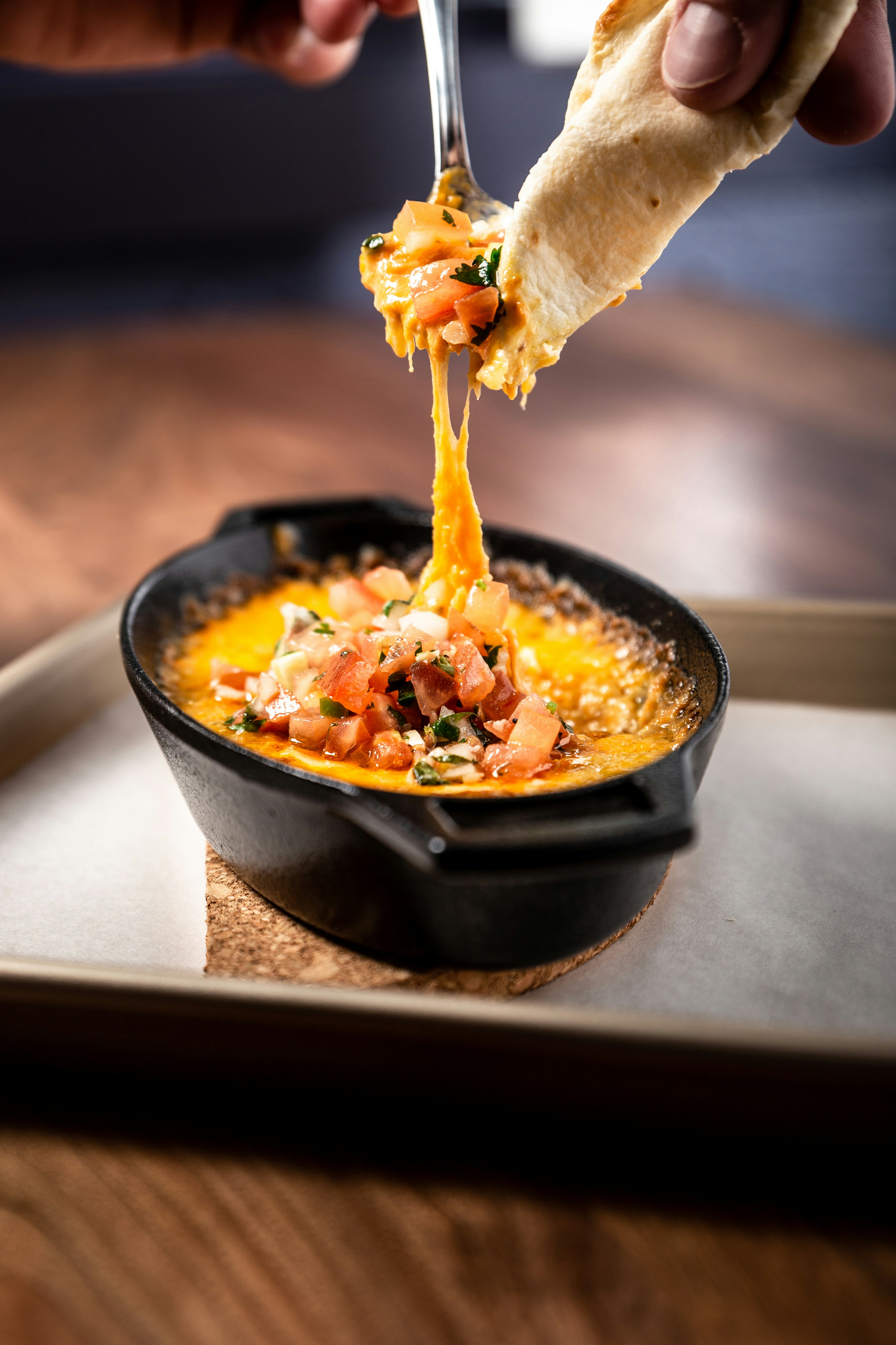 Melted cheese topped with fresh tomatoes and herbs, served in a cast iron dish. A hand reaches in with a tortilla for a delicious dip.