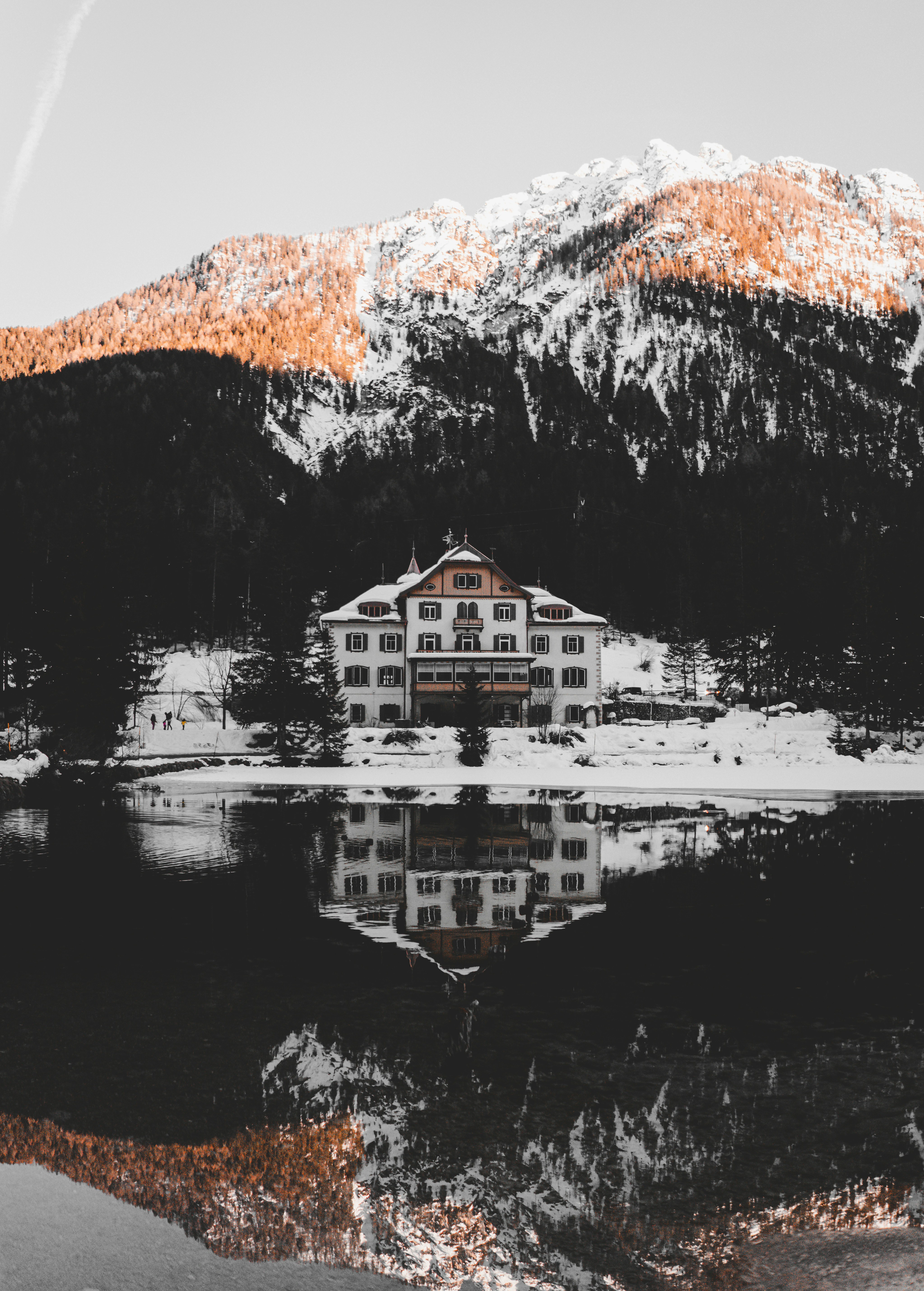 Historic lodge nestled by a snow-covered lake, reflecting the surrounding mountains and trees. Tranquil winter scene capturing nature's beauty.
