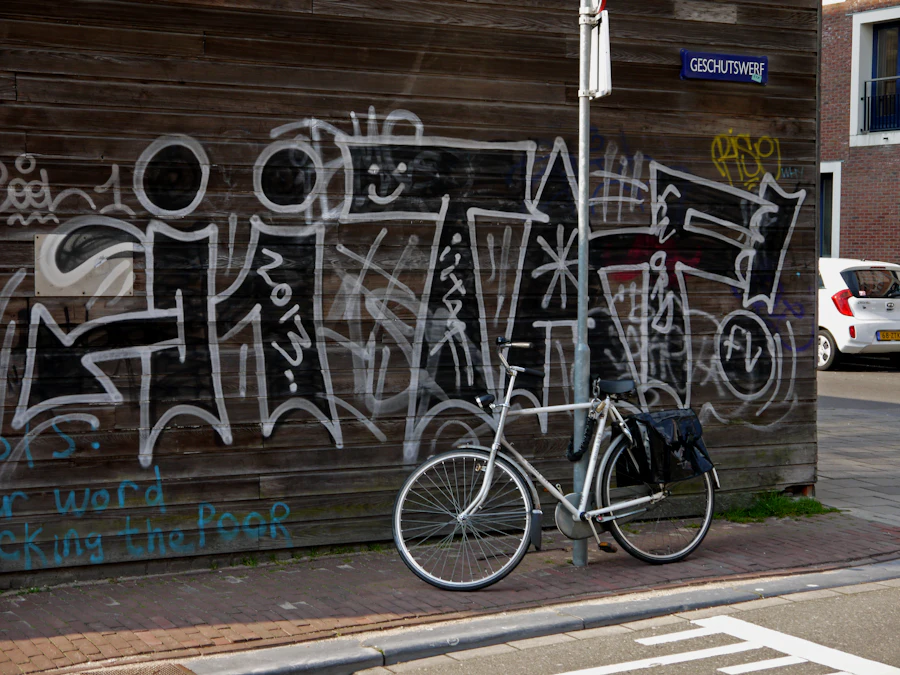 A silver bicycle is locked to a pole in front of a graffiti-covered wooden wall on a city street. A parked car peeks in from the right edge.