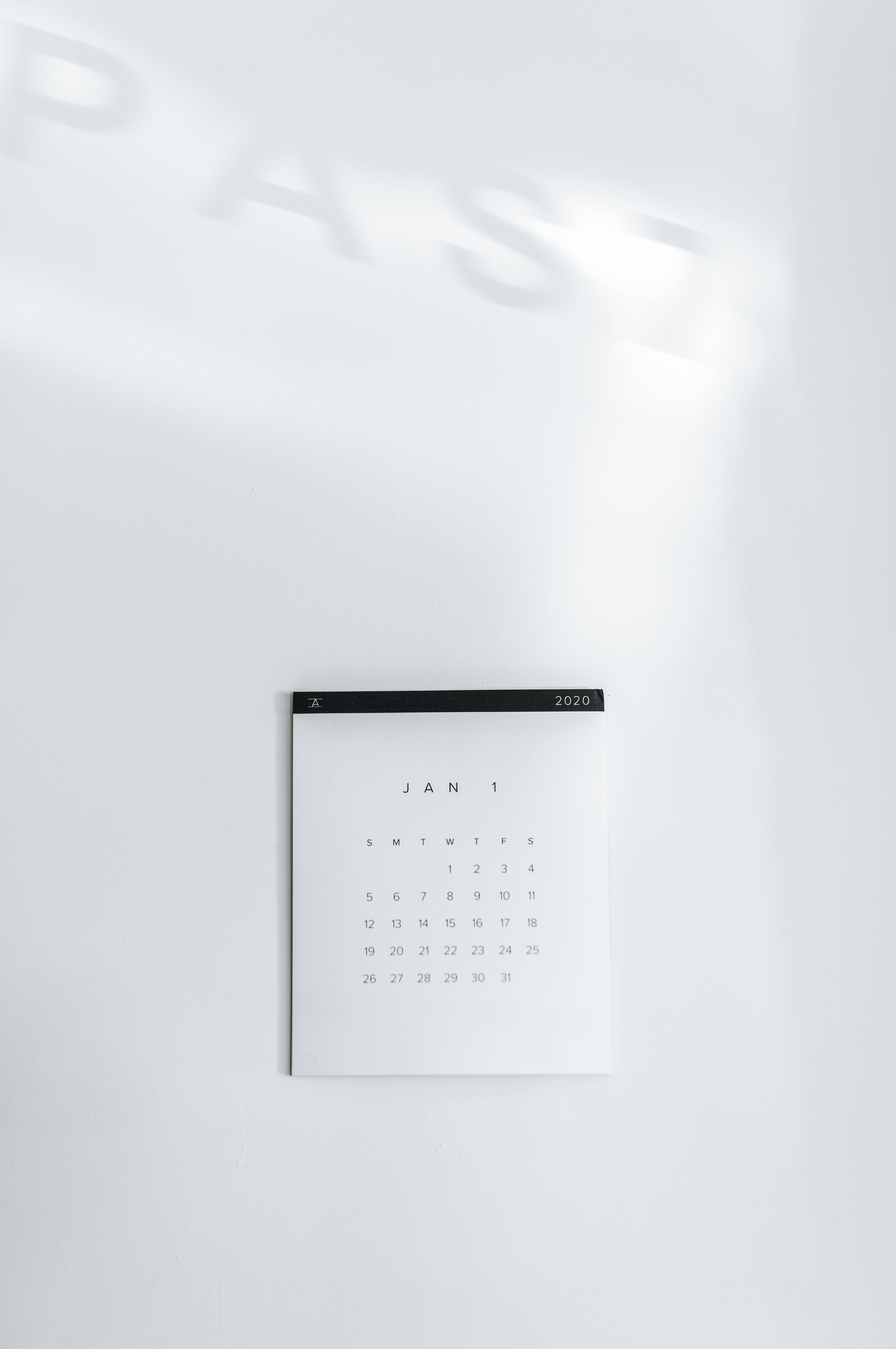 Minimalist calendar displaying January 1, 2020, against a softly lit white wall, with the word 'PAST' subtly cast in shadow.