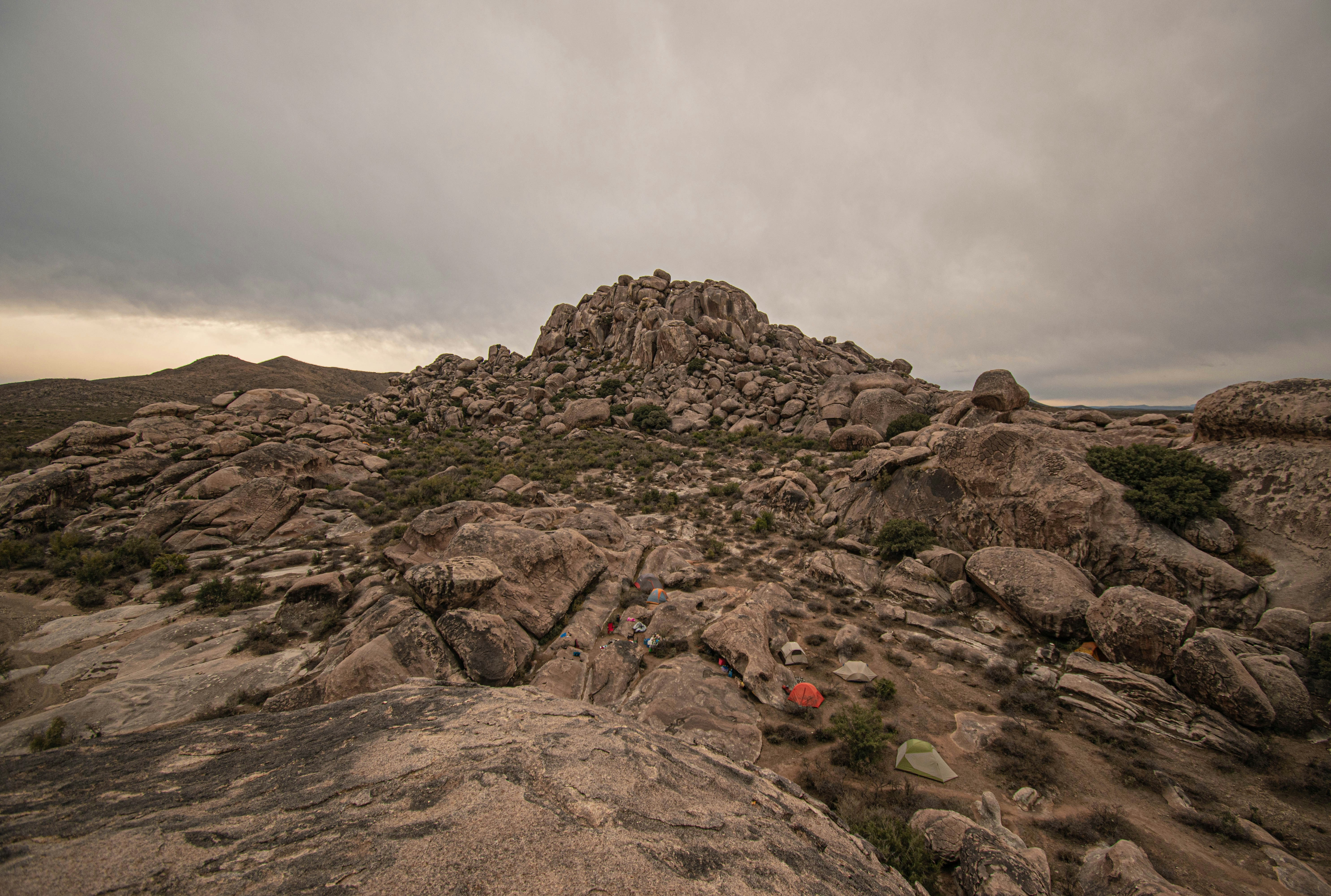 A rugged landscape featuring large boulders and sparse vegetation under a cloudy sky, with hints of camping gear visible in the foreground.