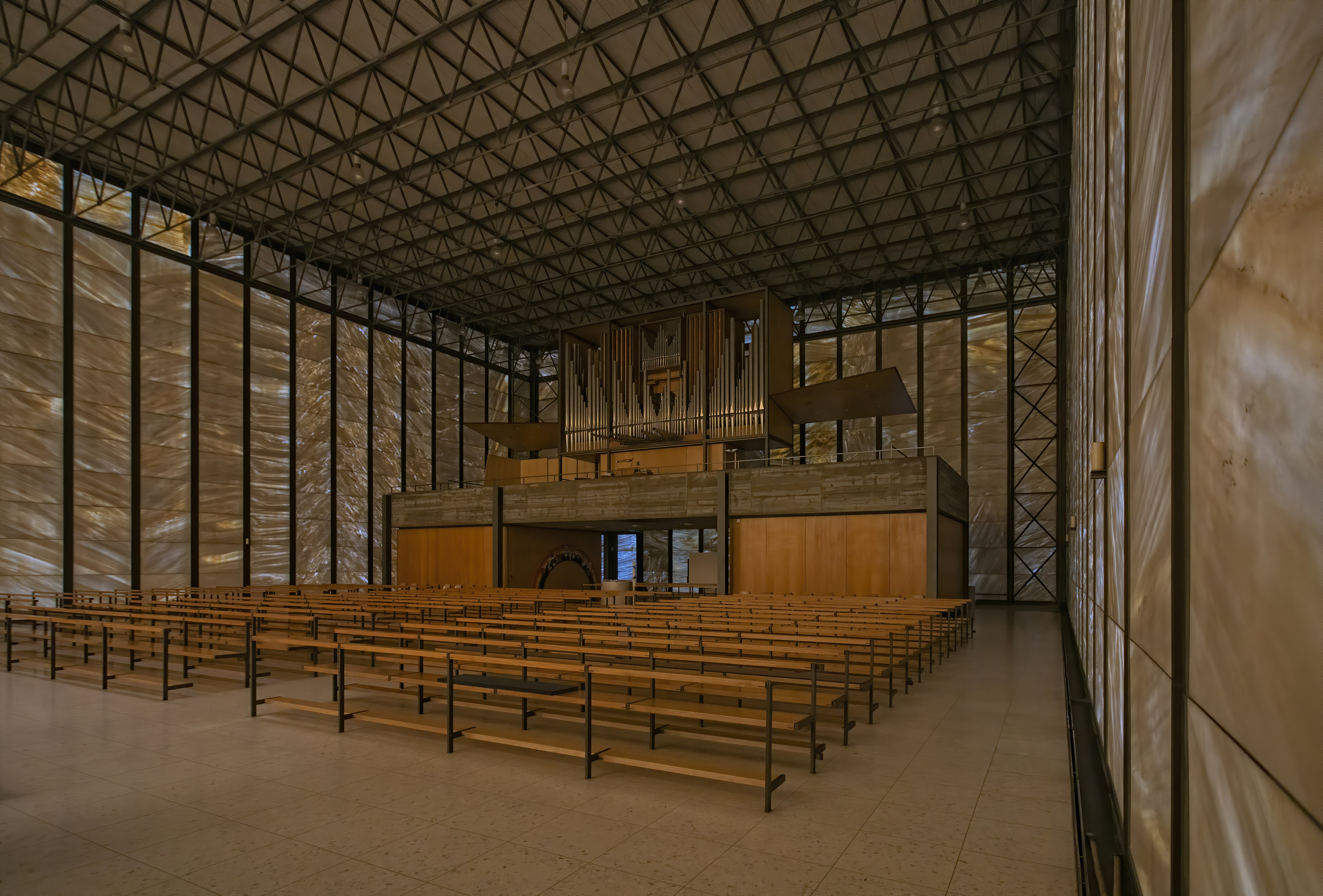 Interior of a modern chapel featuring a prominent organ and minimalist wooden benches, illuminated by natural light filtering through large windows.