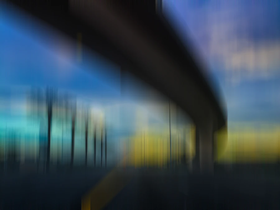 Abstract motion-blurred scene of an elevated roadway cutting across a blue and yellow twilight sky, with dark vertical structures below.