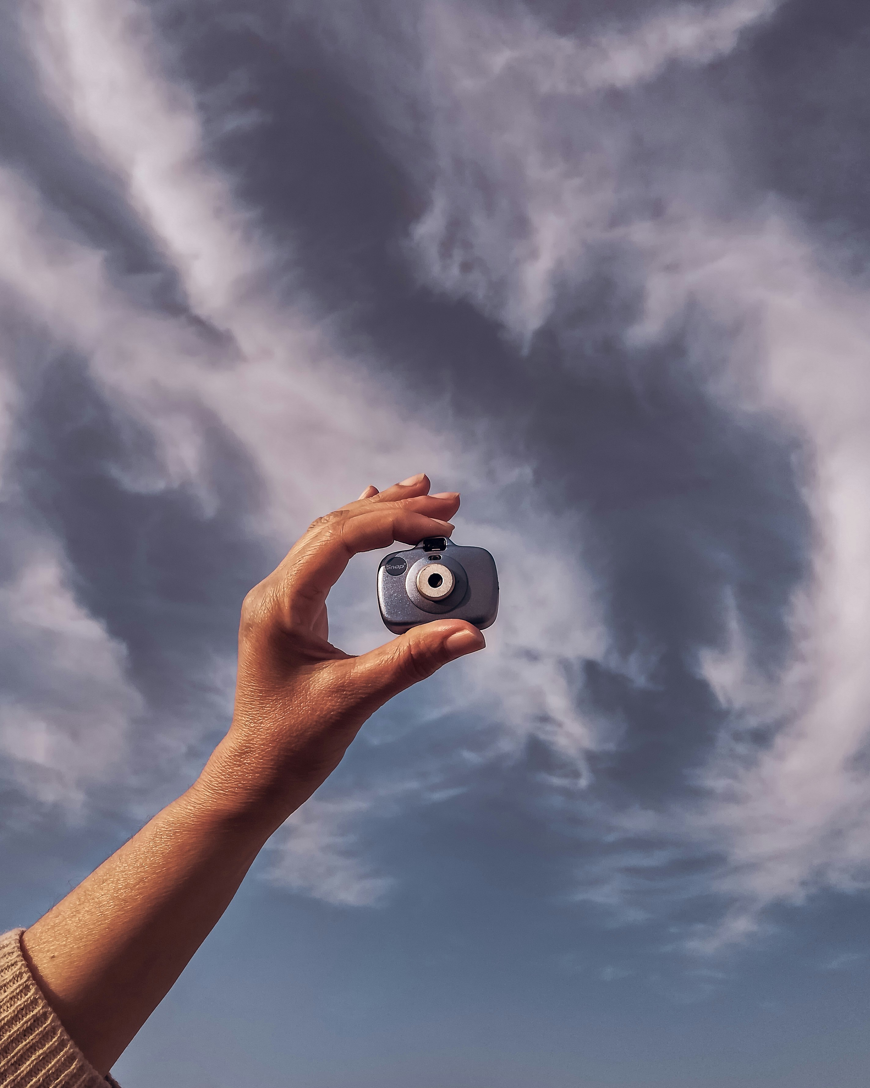 Hand holding a tiny camera against a backdrop of wispy clouds in a blue sky.