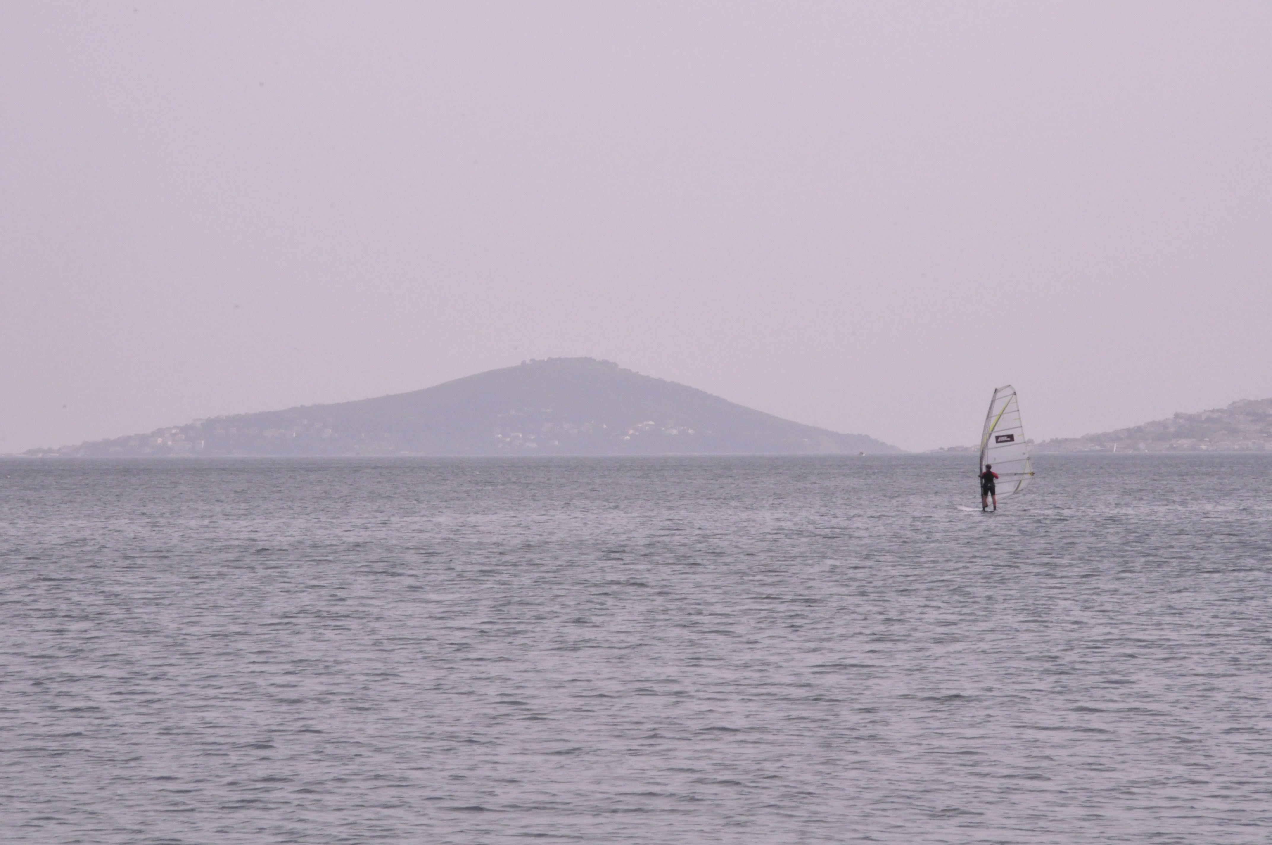 Pastel Wake: A lone windsurfer threads a quiet seam of sea toward a distant island