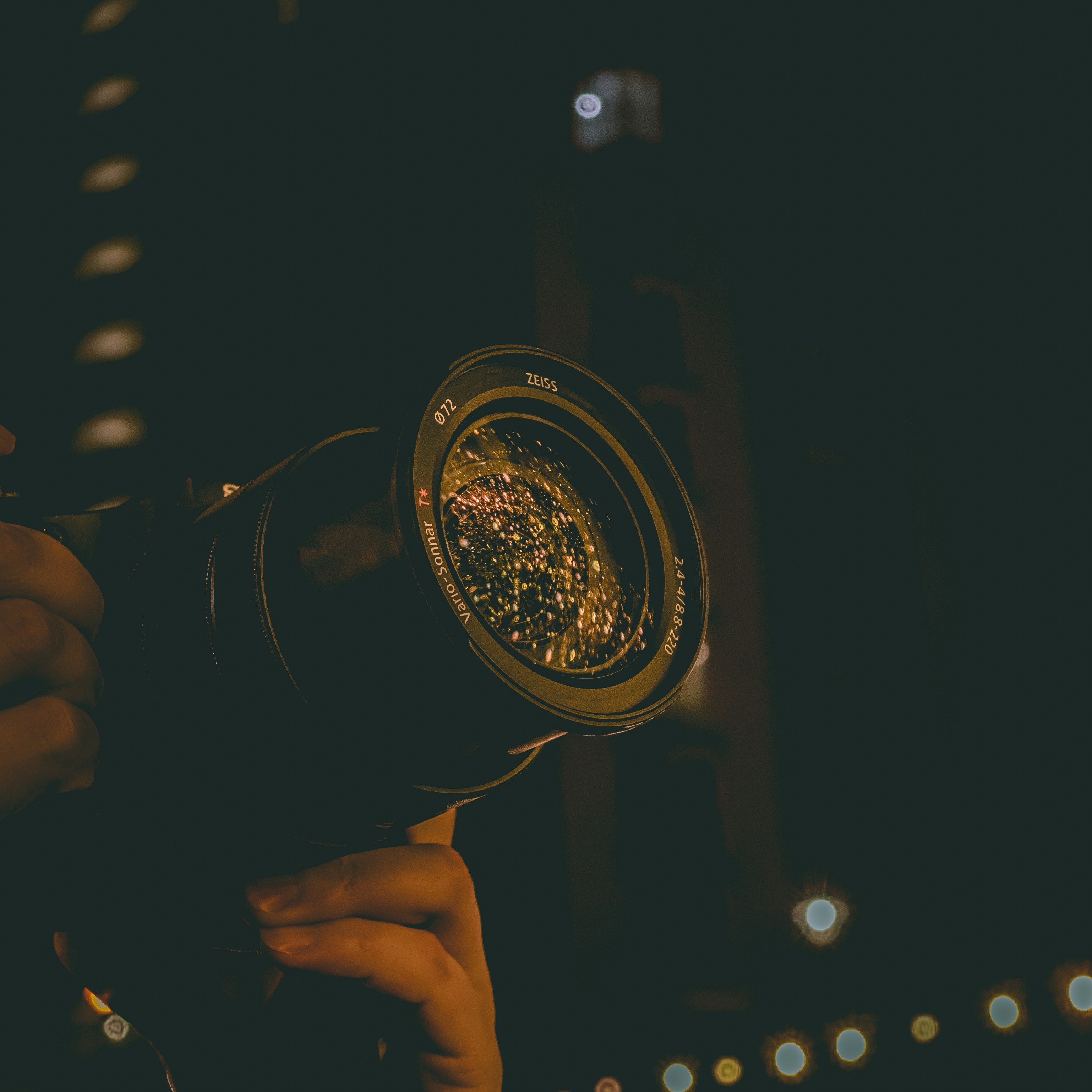 Photographer holds a compact camera with a large lens in a dark urban setting. Distant streetlights blur into warm bokeh, highlighting the gear and mood.