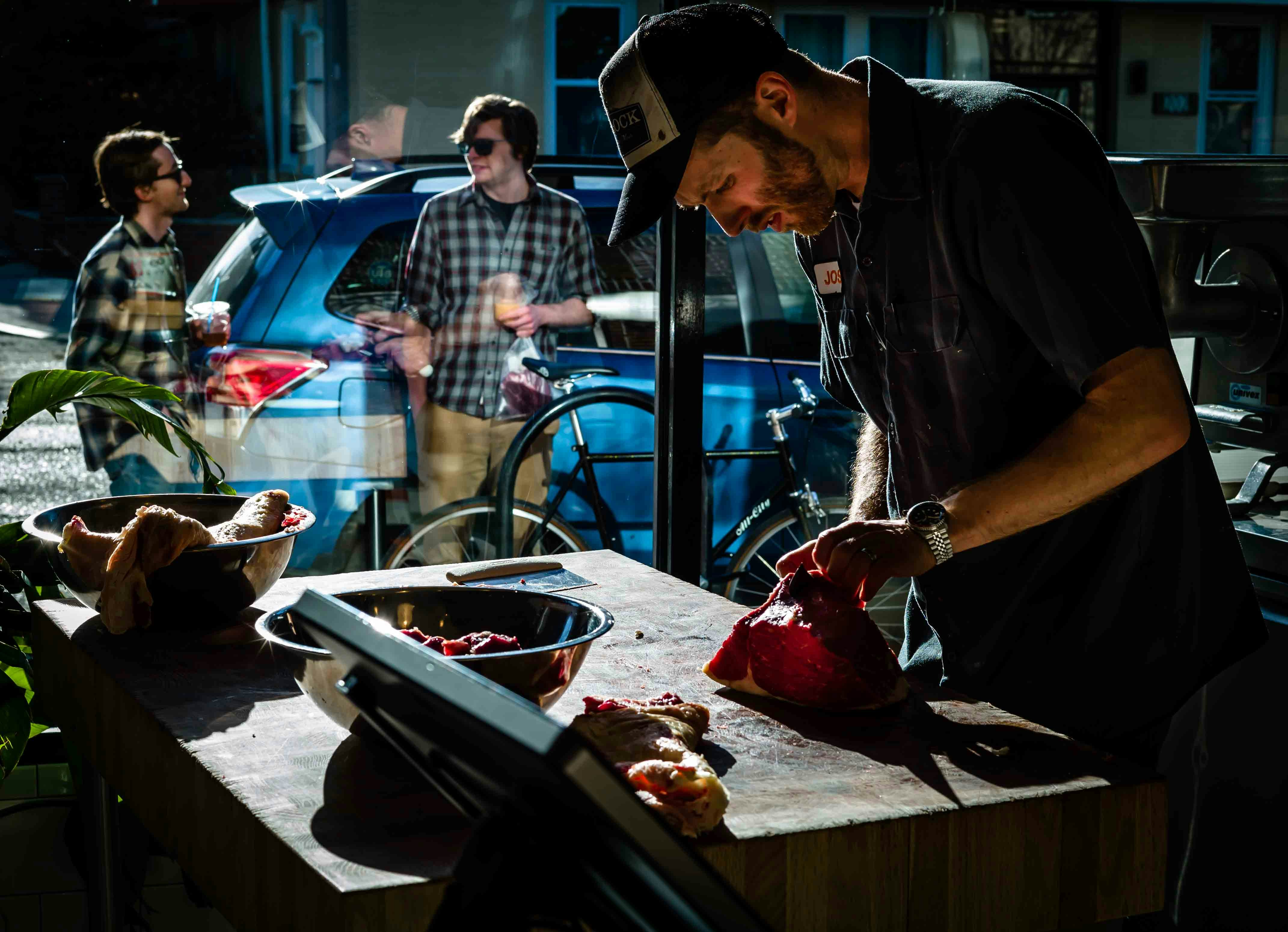 Chef slicing meat at a wooden counter with a bowl of meat slices, sunlight streaming through a window.