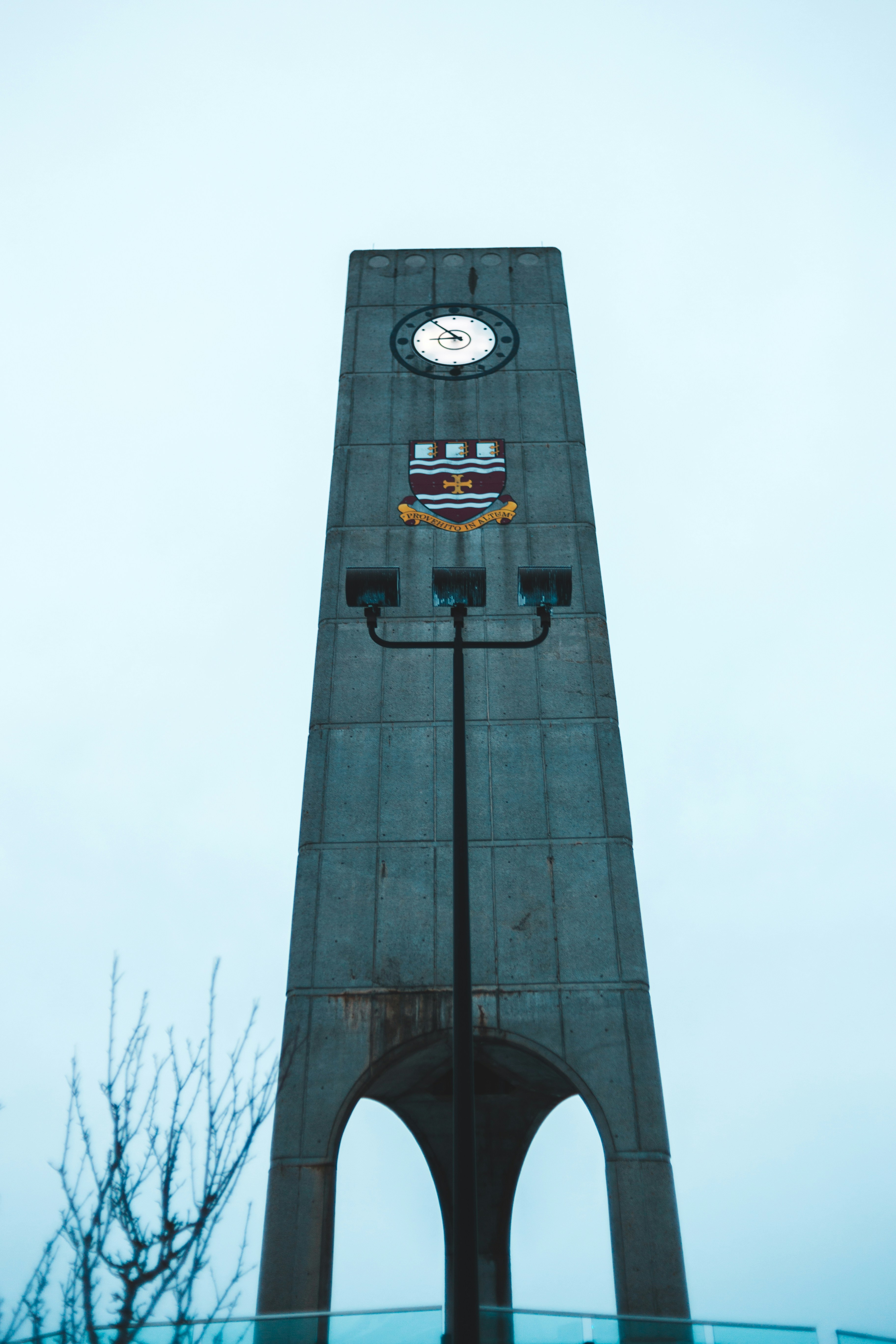 Concrete clock tower adorned with a university crest, standing tall against a pale sky. The structure features a prominent clock face and streetlight, emphasizing its role as a landmark.