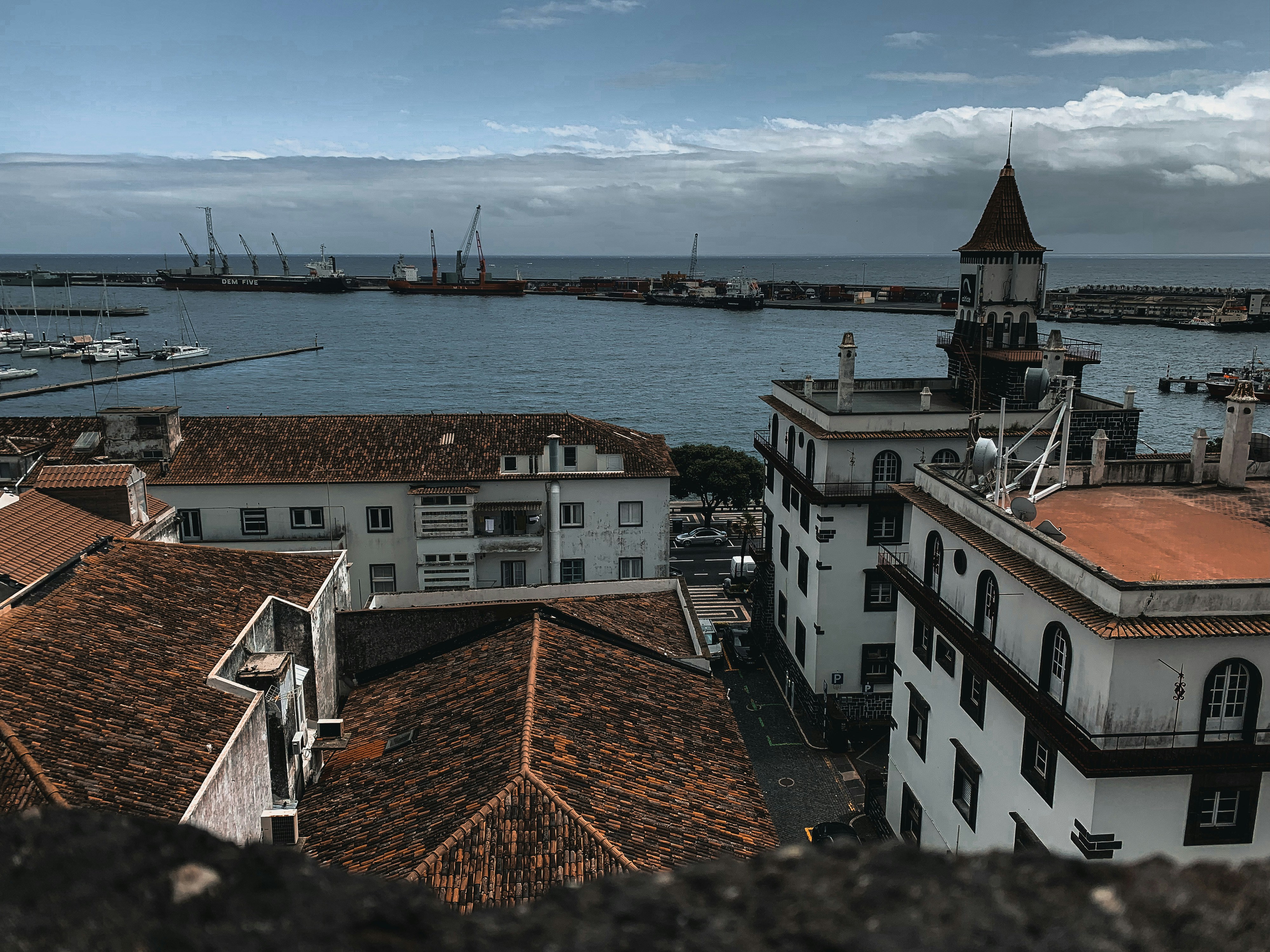 A panoramic view of a coastal harbor, showcasing terracotta rooftops and a distant marina with ships docked. The scene is framed by a mix of historic and modern architecture.