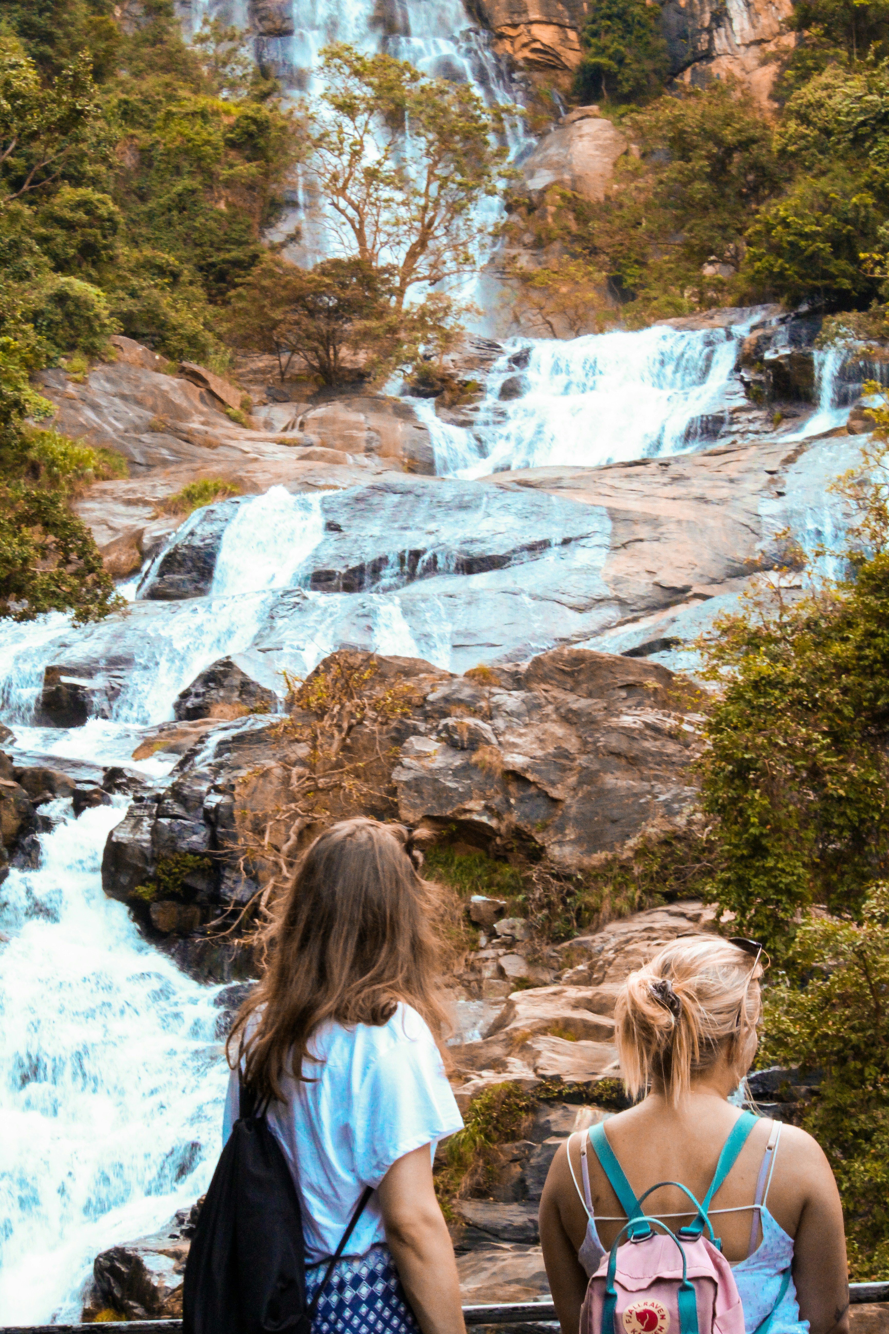 Two individuals admire a majestic waterfall cascading down rocky terrain, surrounded by lush greenery.