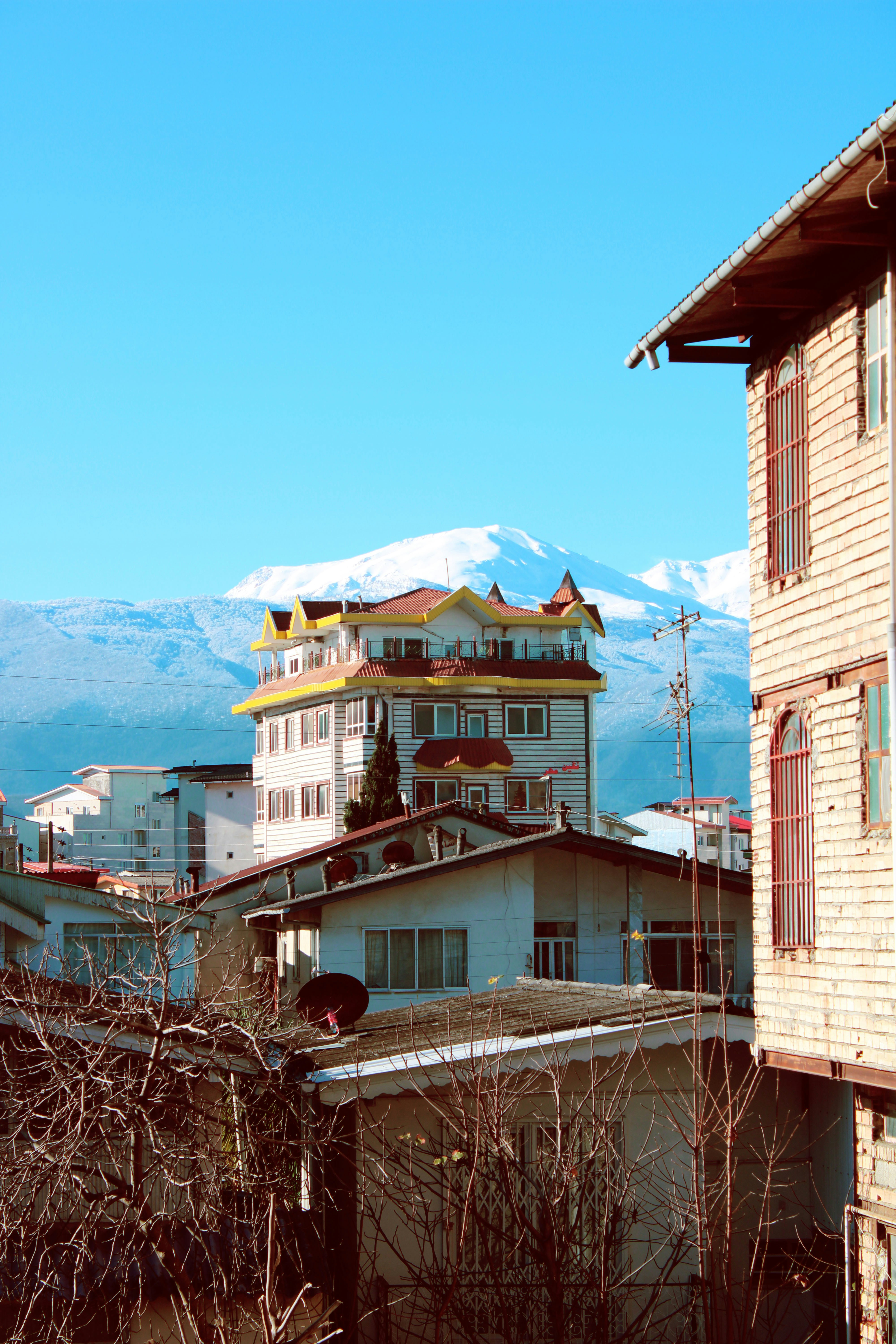 A picturesque townscape featuring a prominent multi-story building framed by rustic homes, with majestic snow-capped mountains in the background. The clear blue sky enhances the tranquil atmosphere.