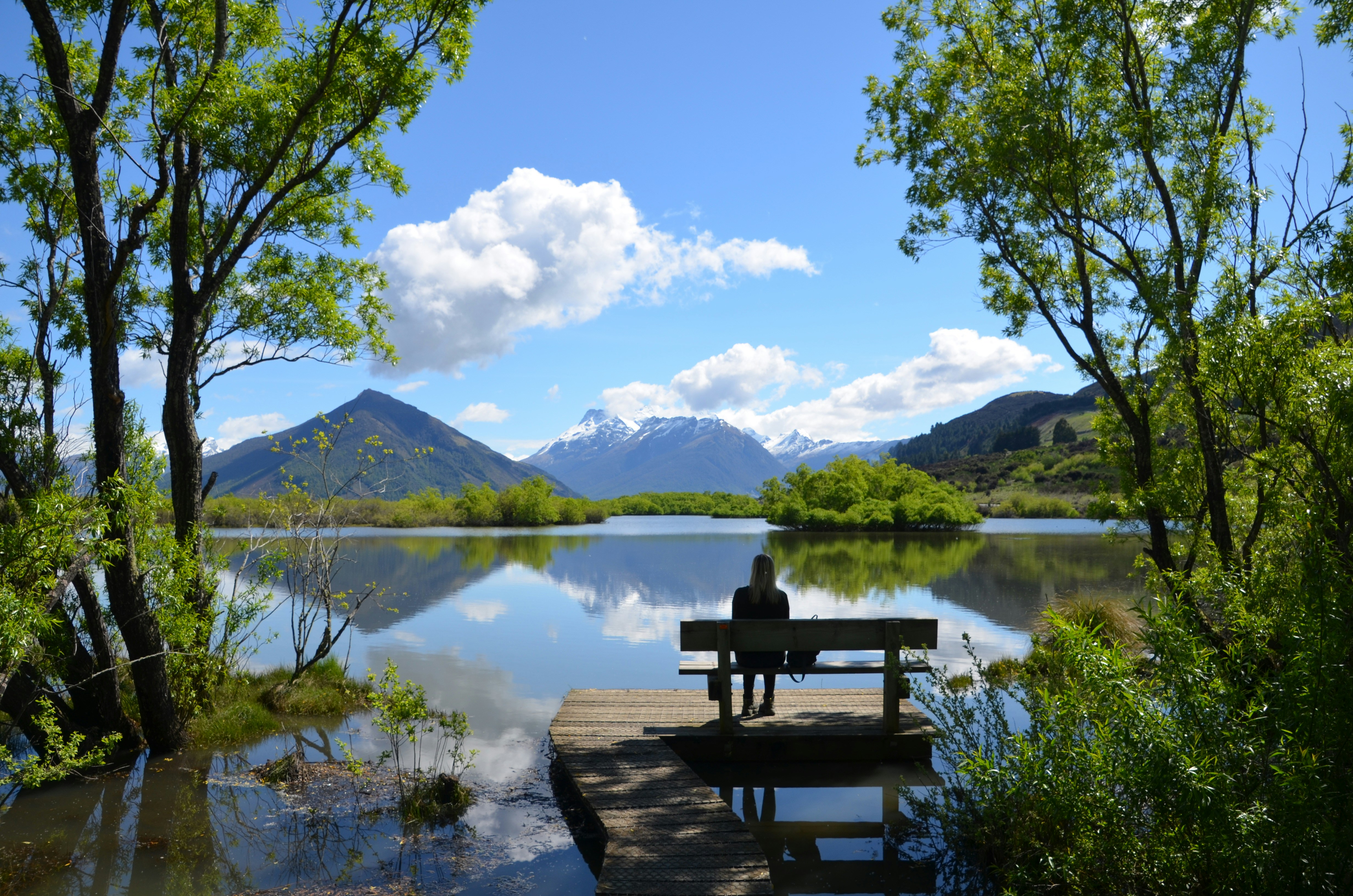 Moments before I dropped my phone in the water. Description: Glenorchy lagoon in New Zealand with a small boardwalk, some trees to the side, a seat with a person in it overlooking the lagoon, and mountains in the background reflecting off of the lake. | woman sitting on bench near lake