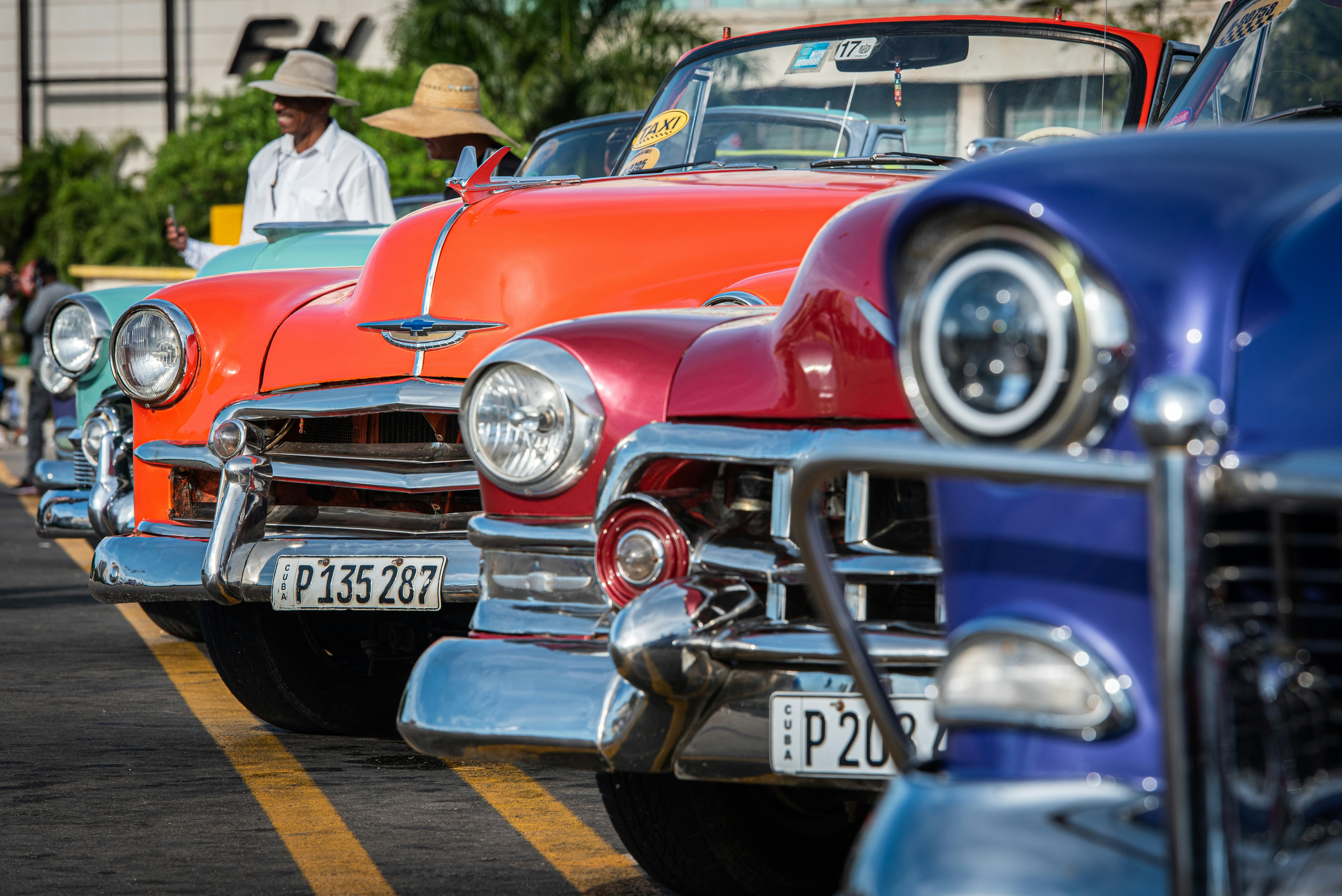 This captivating image showcases a lineup of classic vintage cars in Havana, with a focus on their vibrant colors including bright orange, red, and blue. The composition highlights the gleaming chrome details and distinctive designs of the vehicles, set against a lively street scene. The image captures the essence of nostalgia and cultural richness, enhanced by bright daylight that accentuates the vivid hues and polished surfaces.