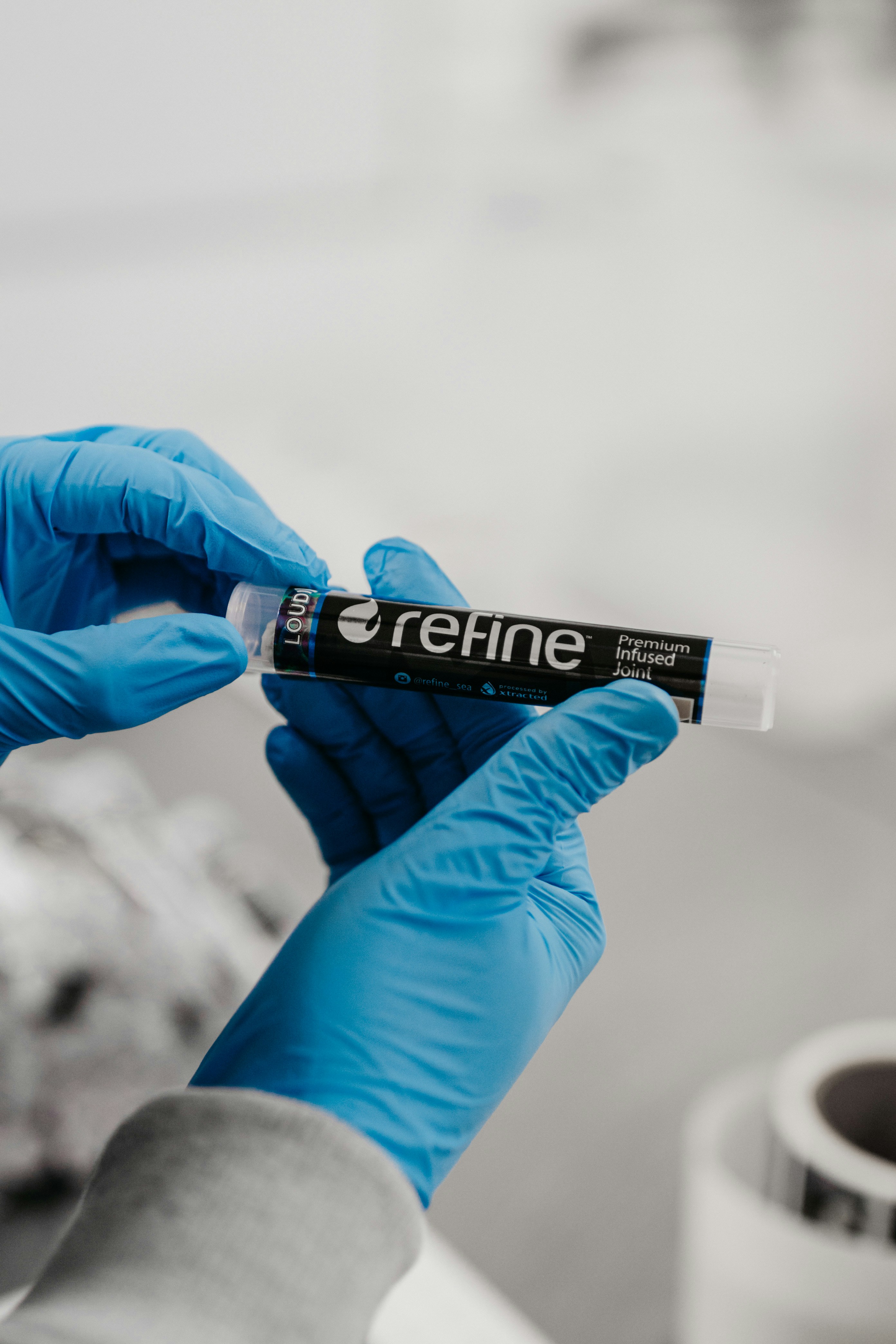 Blue-gloved hands hold a sleek droplet of gel labeled 'refine' in a laboratory setting