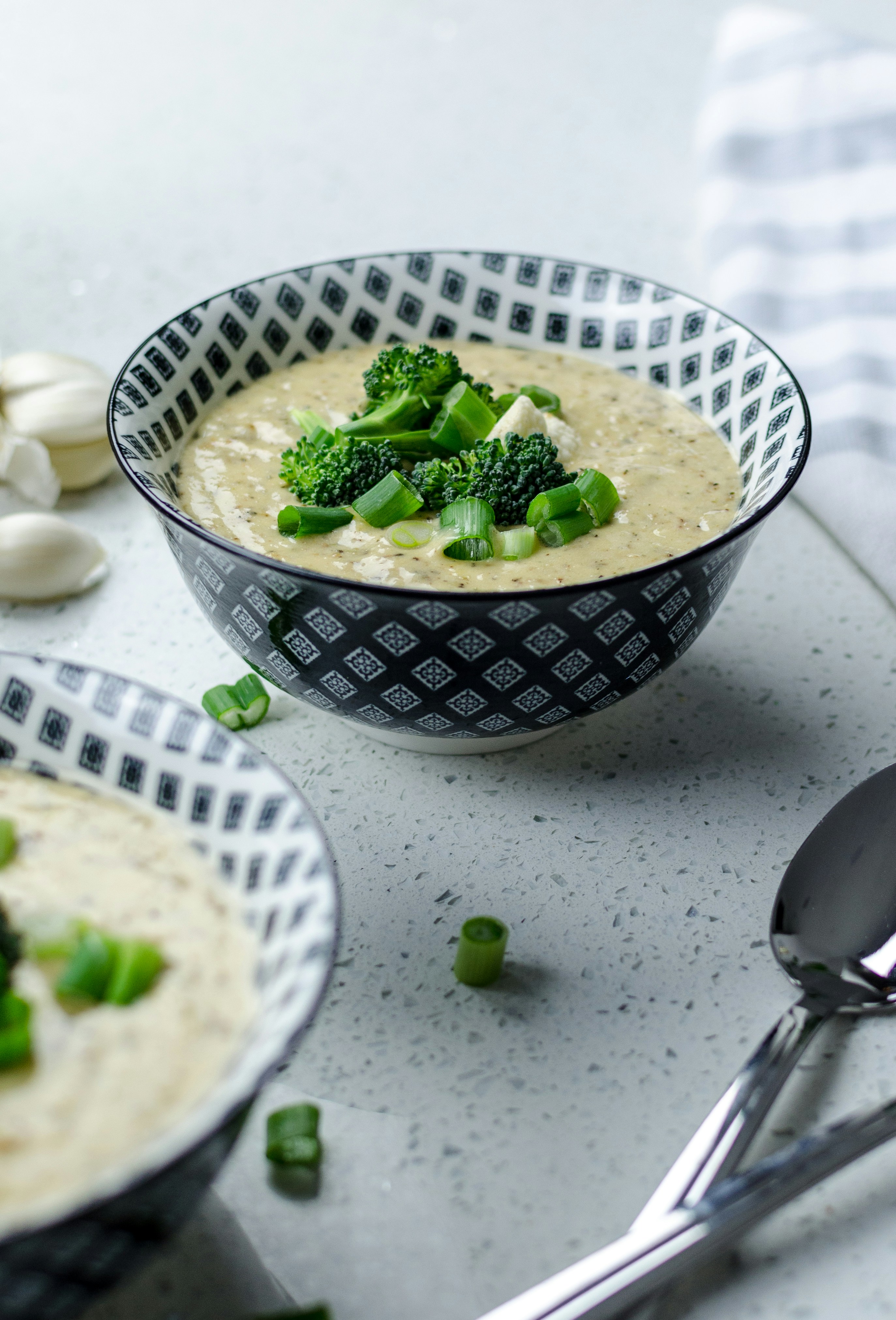 A bowl of creamy soup garnished with broccoli and green onions, set against a minimalist countertop. The intricate bowl design adds a touch of elegance.
