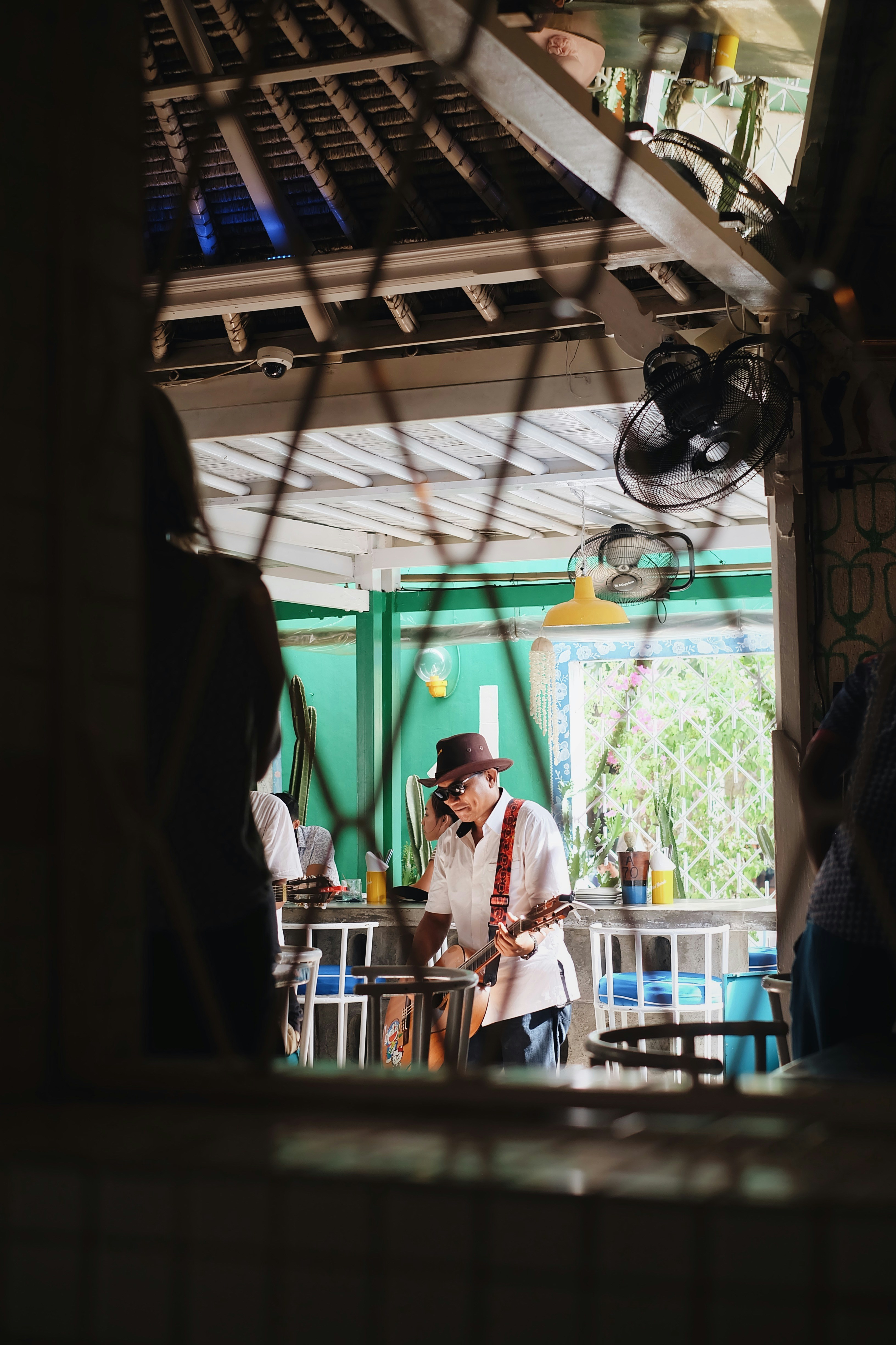 Musician in a black hat plays guitar in a sunlit café corner, surrounded by vibrant decor and patrons.
