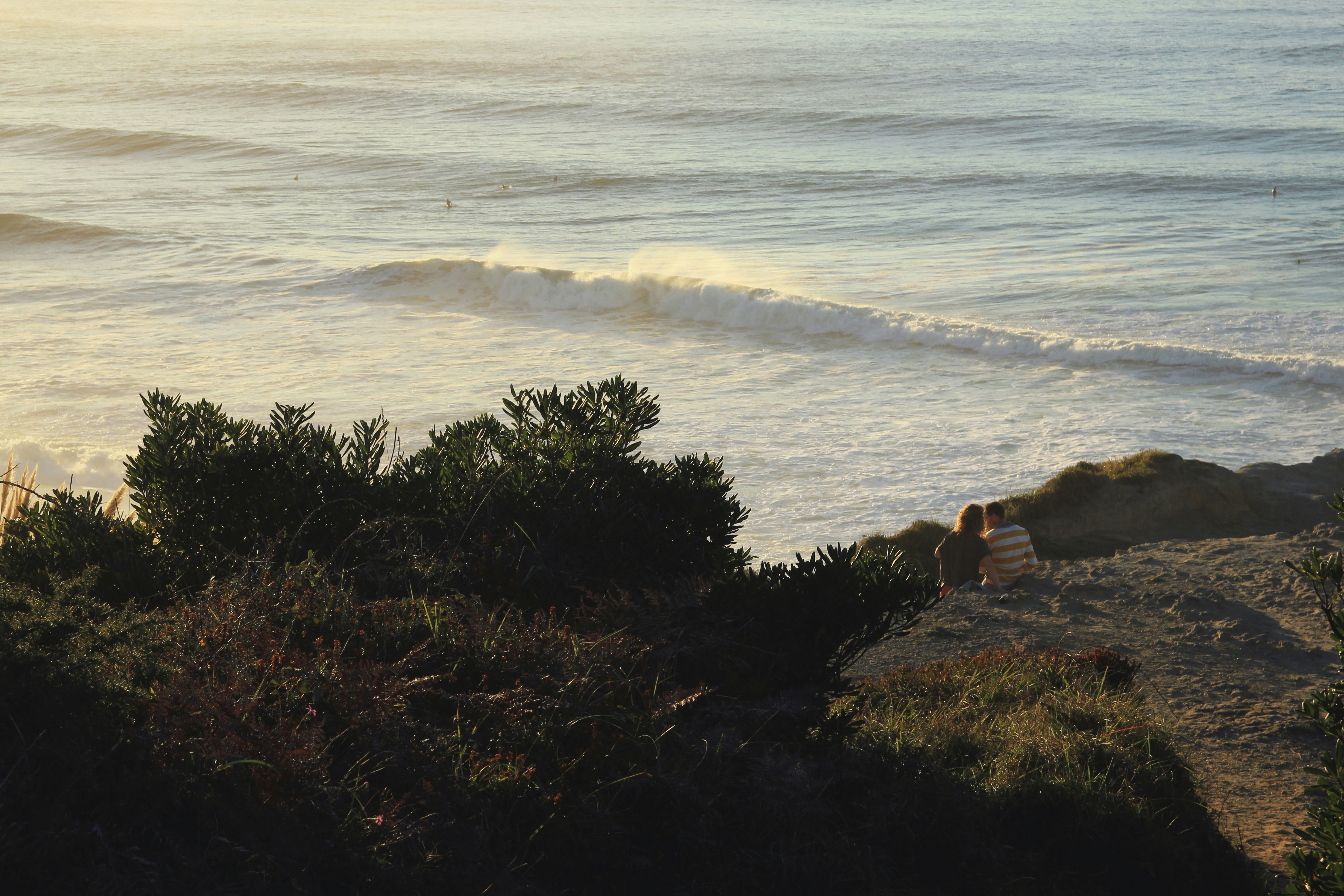 Couple sitting on a cliffside, gazing at gentle waves rolling onto the shore during sunset. Lush greenery frames the scene.