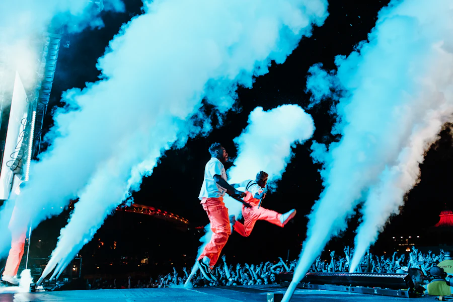 Two performers in bright red pants jump on a concert stage amid towering plumes of blue smoke, with a cheering crowd lit behind them at night.