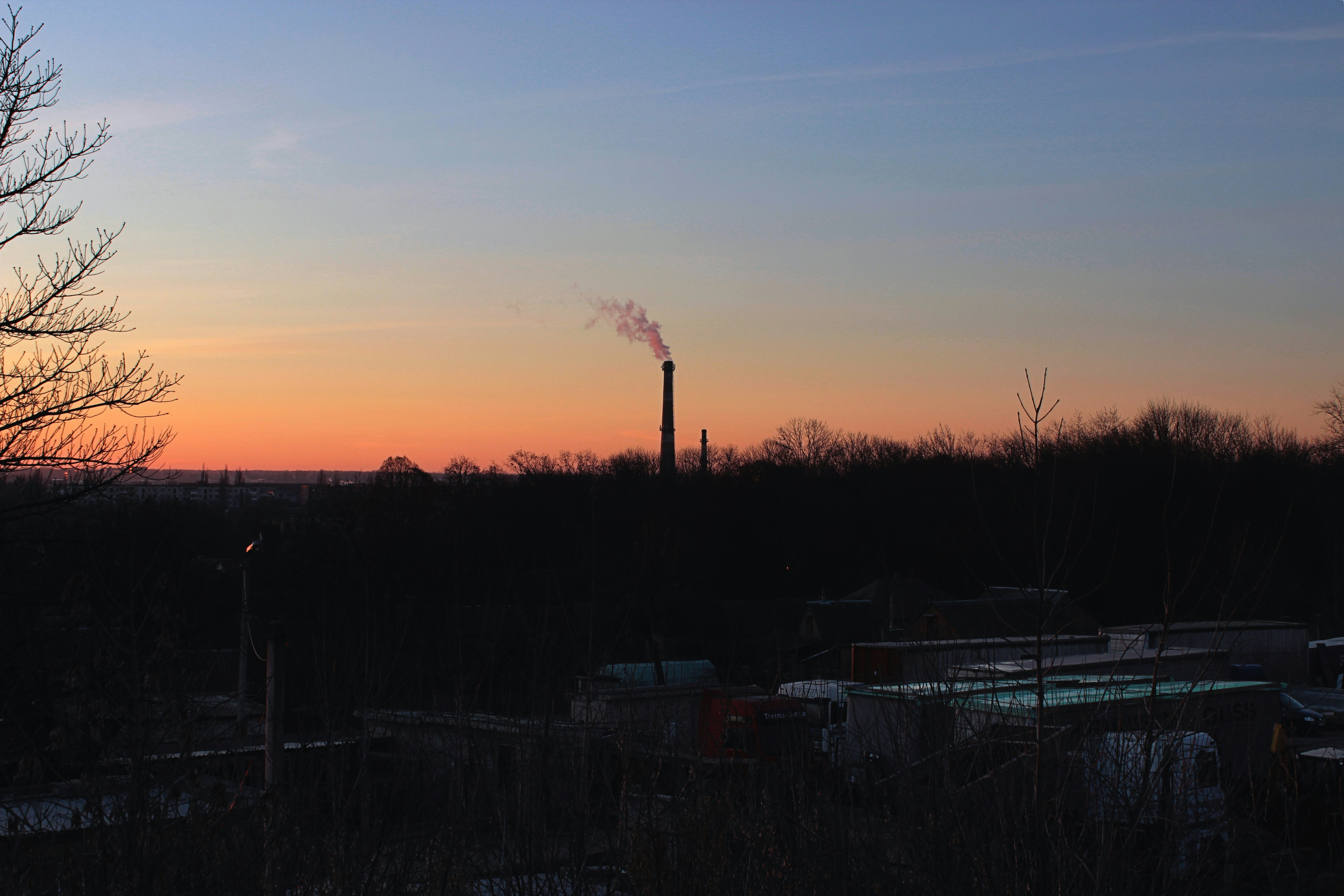 Smoke rises from a solitary factory chimney against a vibrant sunset, silhouetted by the surrounding trees and structures.