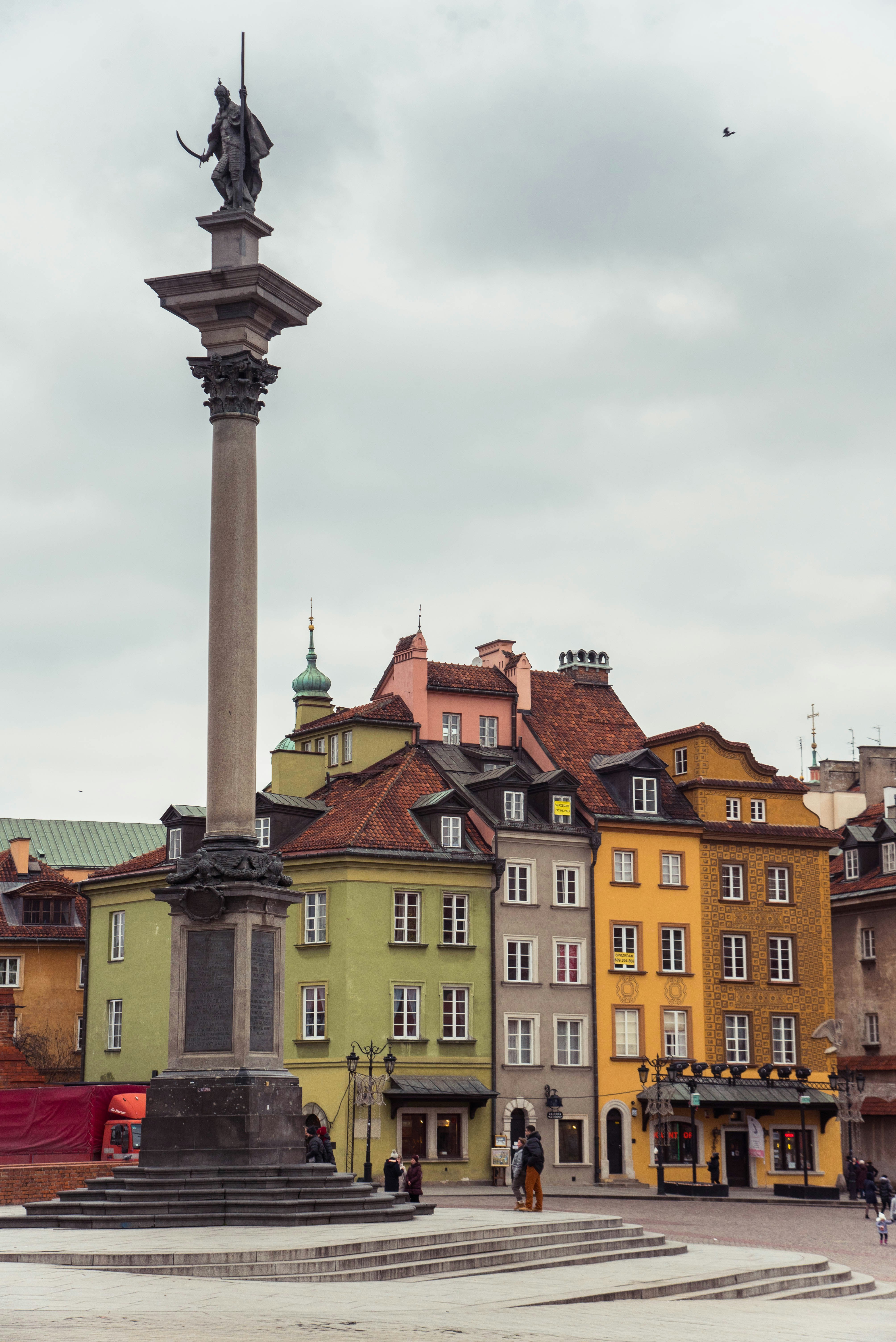 A historic monument stands tall in a vibrant square, surrounded by colorful buildings showcasing architectural diversity. The scene captures the essence of urban life in Warsaw.