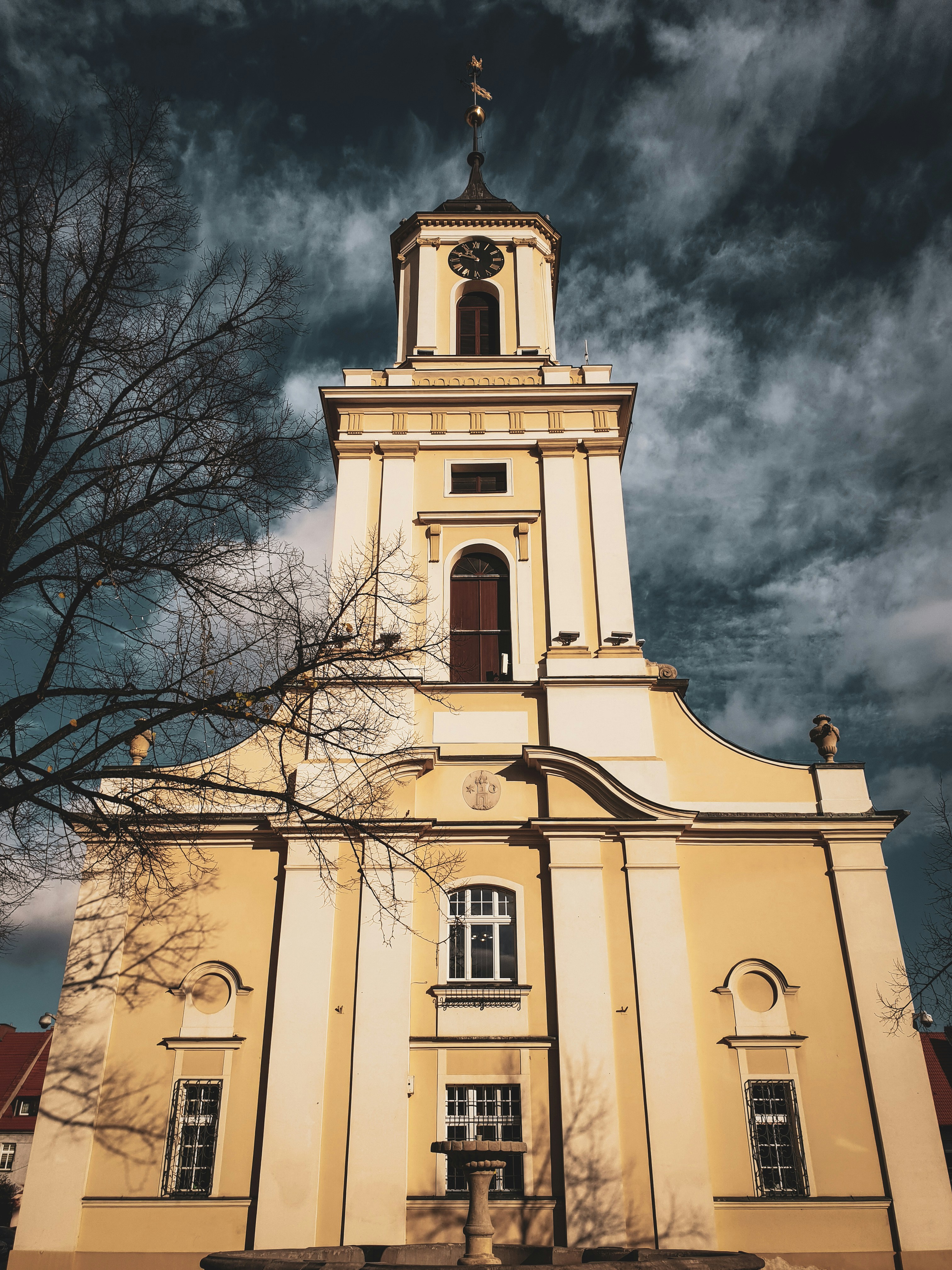 Historic yellow church tower under a dramatic sky, framed by bare branches. The architectural details showcase its grandeur.