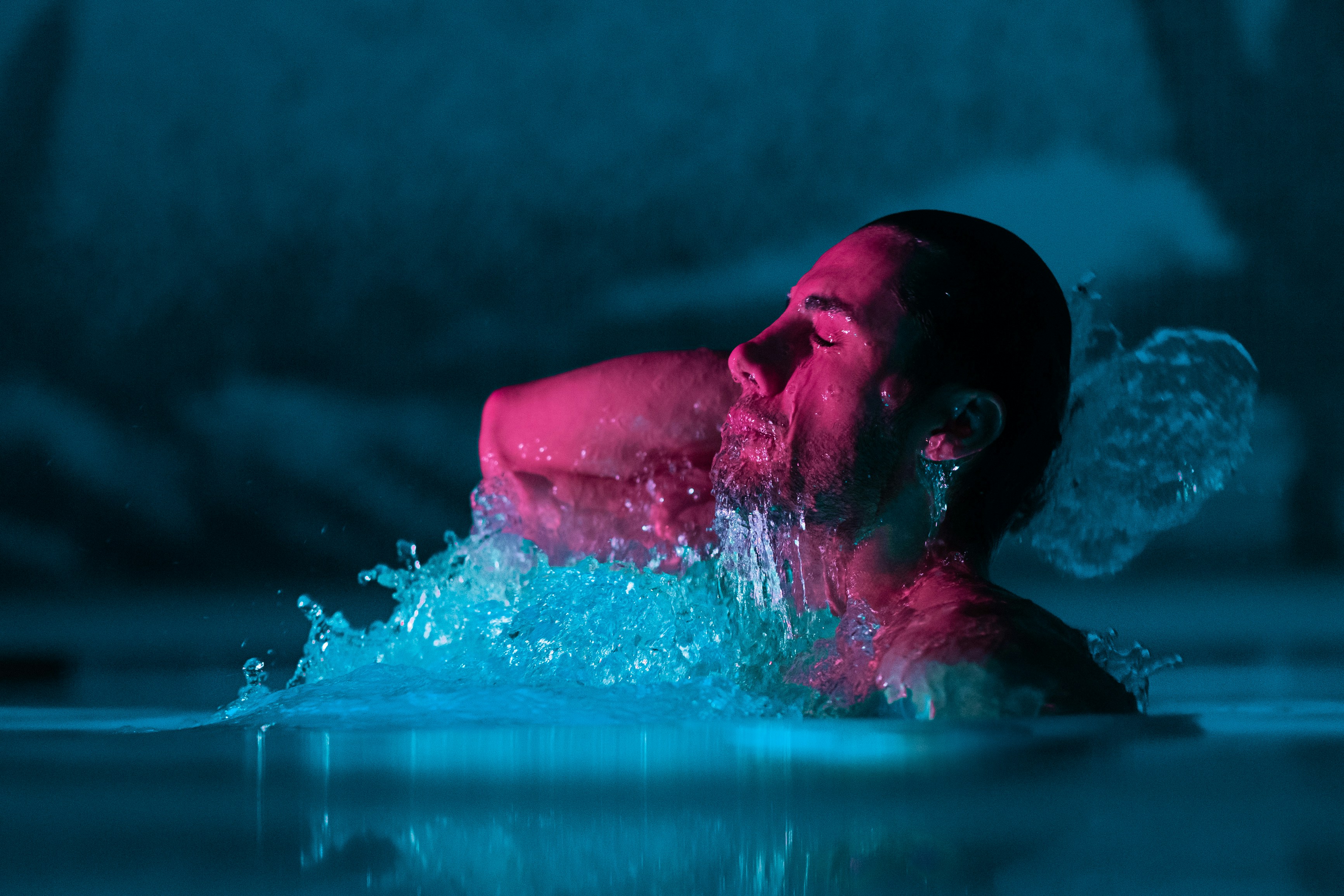 A man emerges from water, illuminated by vibrant blue and pink lighting, creating a dynamic interplay between movement and color.