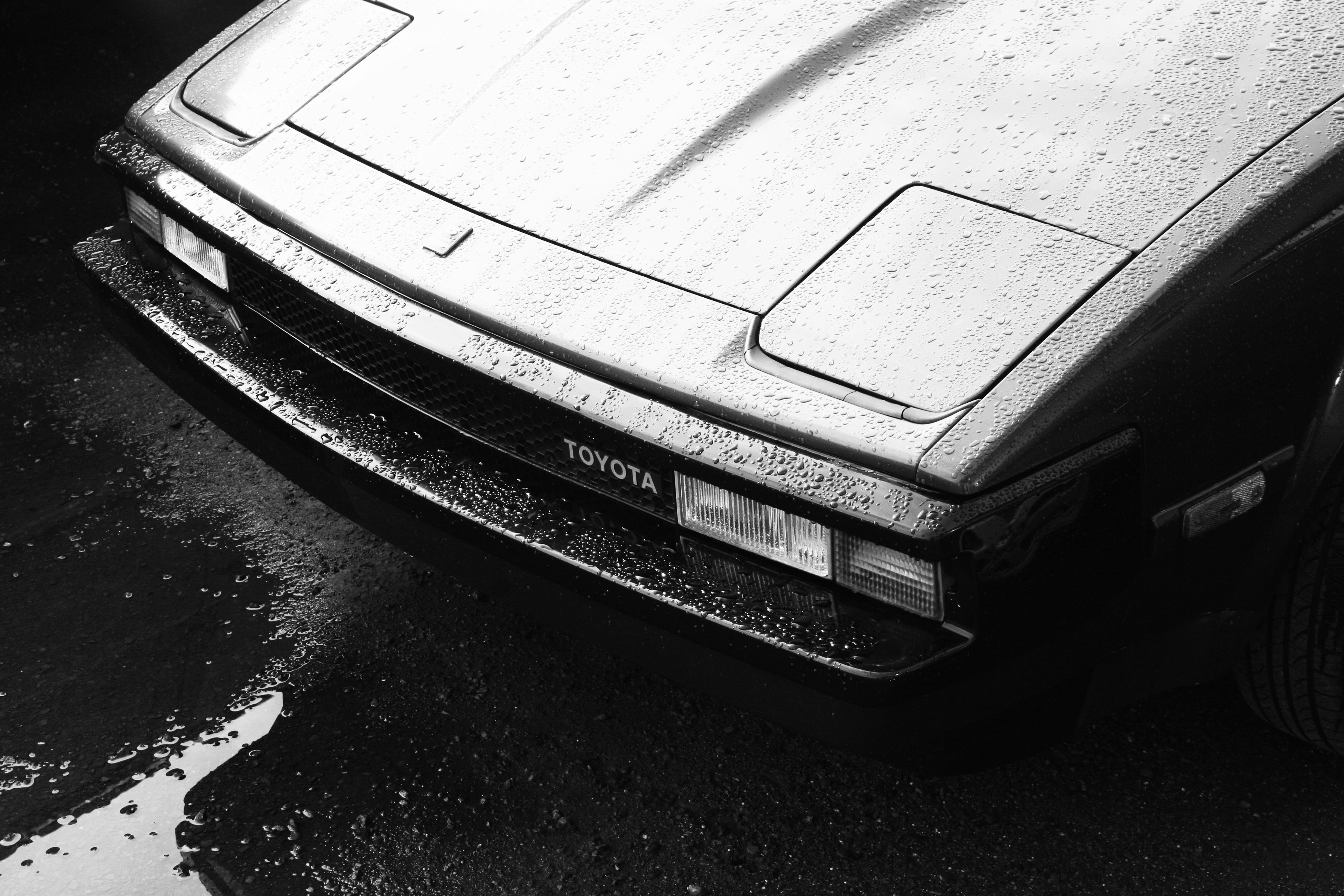 Close-up of a vintage Toyota sports car's front end, showcasing droplets of rain on its sleek black surface.