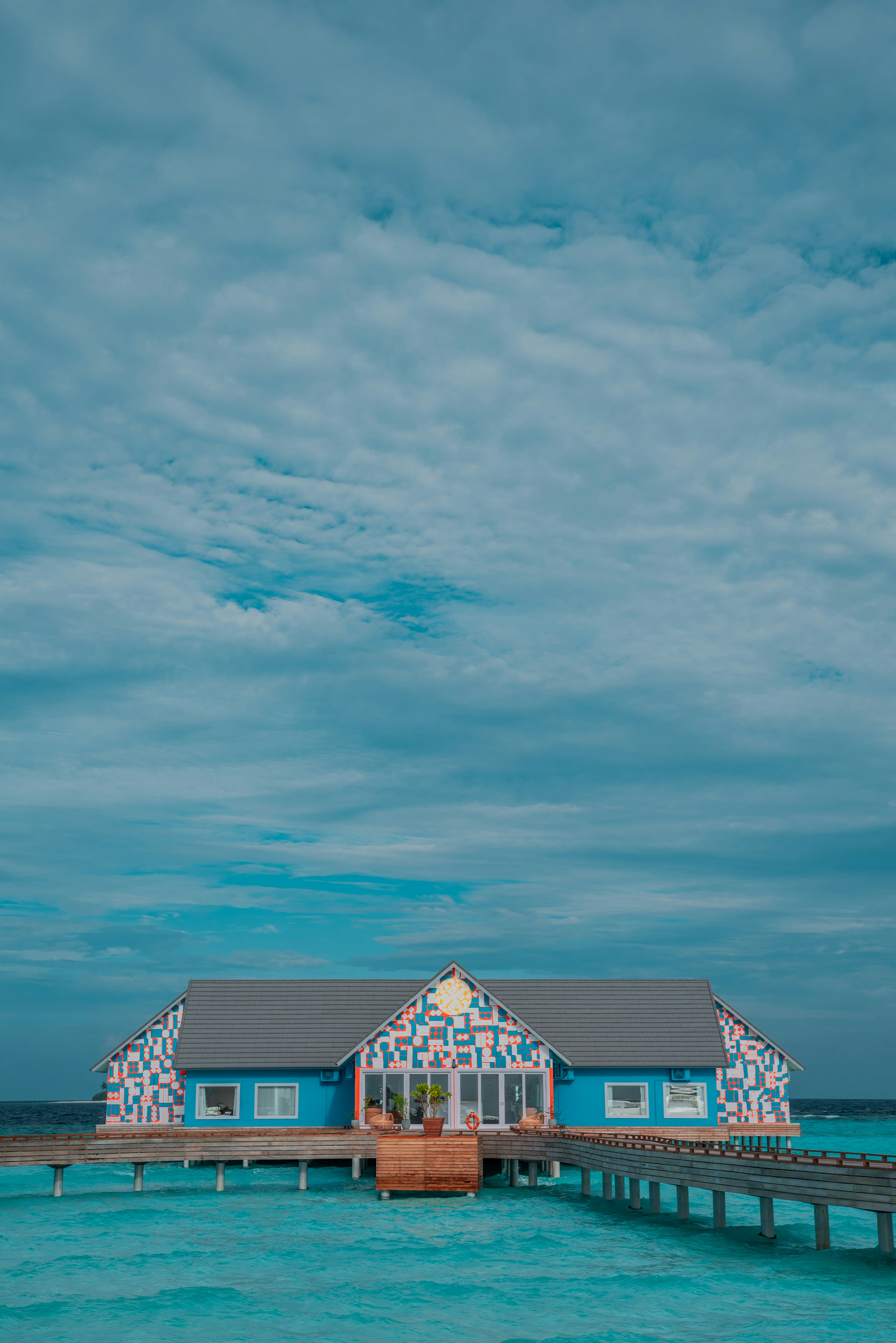 Colorful overwater bungalow with a wooden walkway, surrounded by turquoise waters under a cloudy sky.