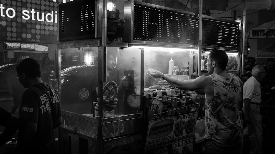 Black-and-white street food stall at night with a man reaching to order from the counter as steam rises under bright lights. Other pedestrians and menu signs frame the busy scene.