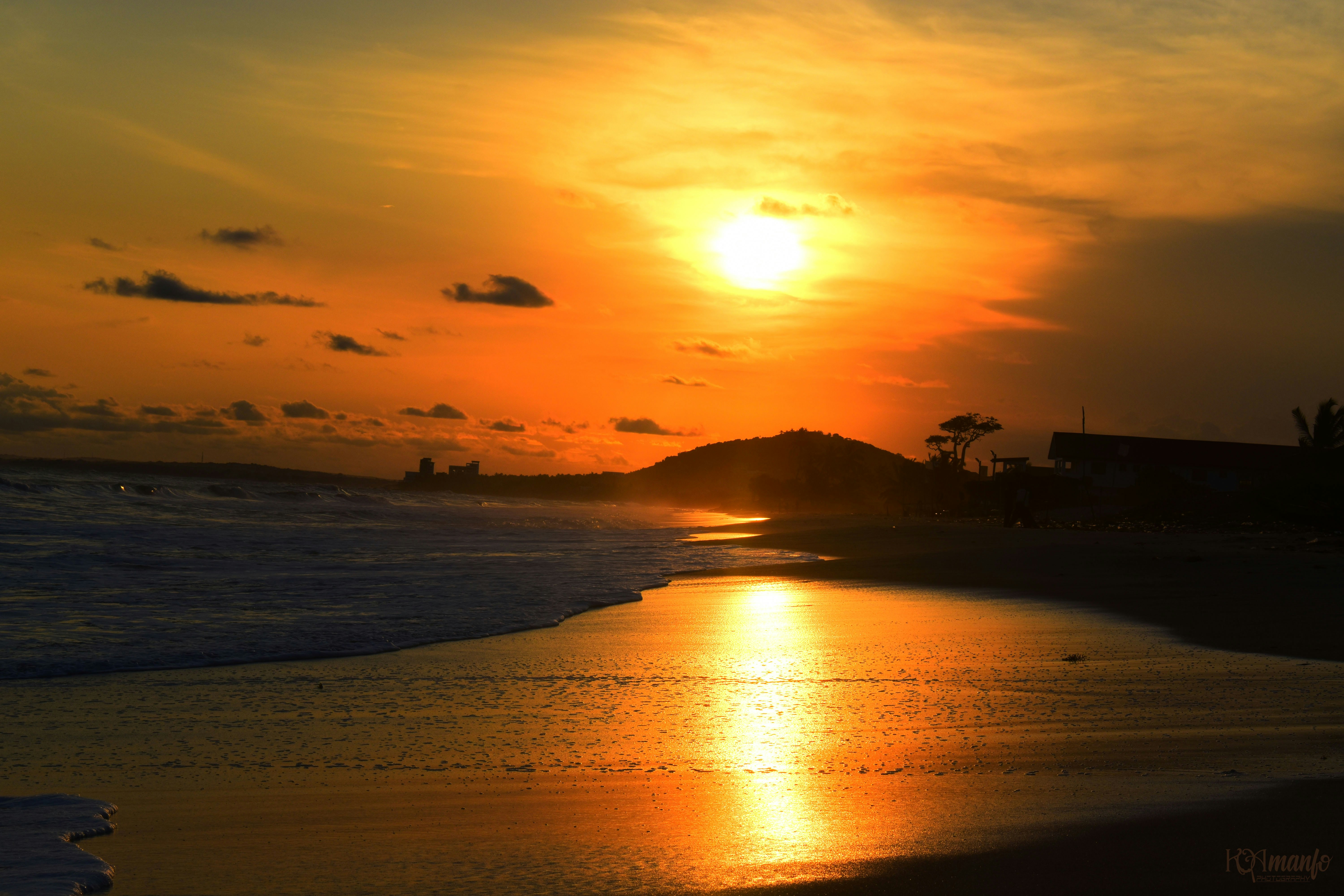 Sunset casting golden hues over a tranquil beach, with gentle waves lapping at the shore and silhouettes of distant hills. 