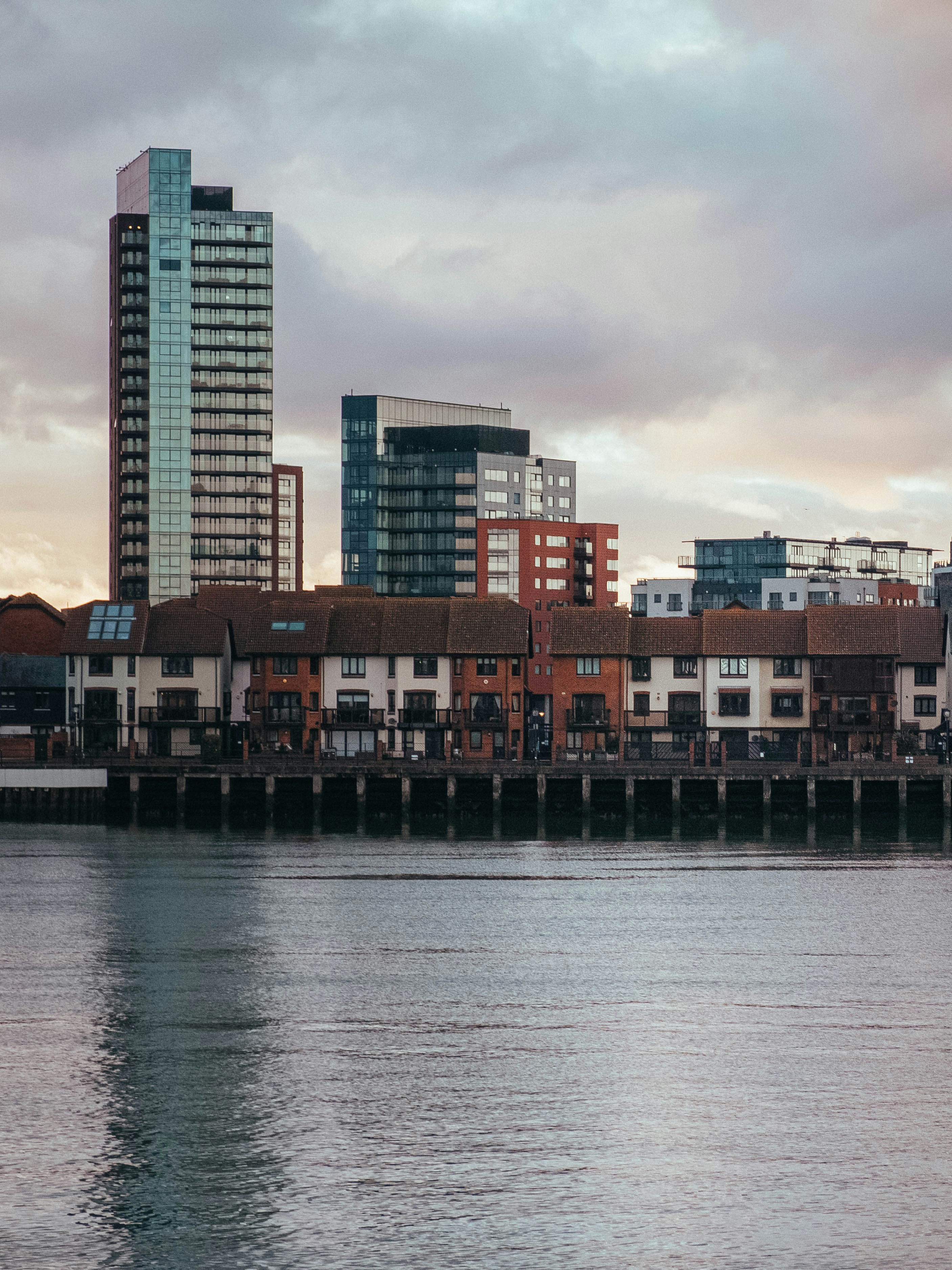 Contemporary buildings lining a waterfront, reflecting in calm waters under a cloudy sky.