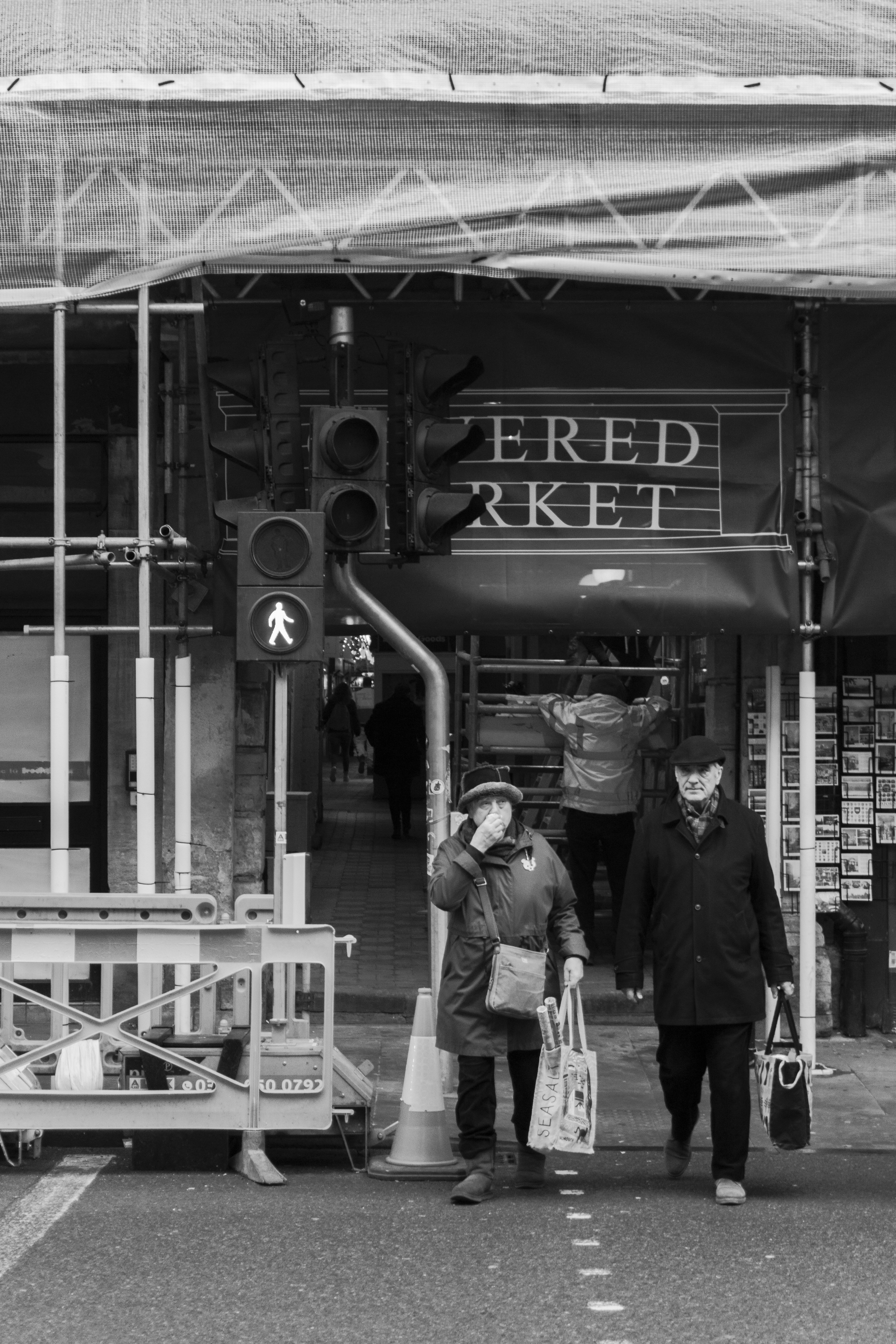 Two pedestrians pause at a traffic signal near a market under construction, embodying the rhythm of city life. The monochrome palette highlights the contrast between movement and stillness.