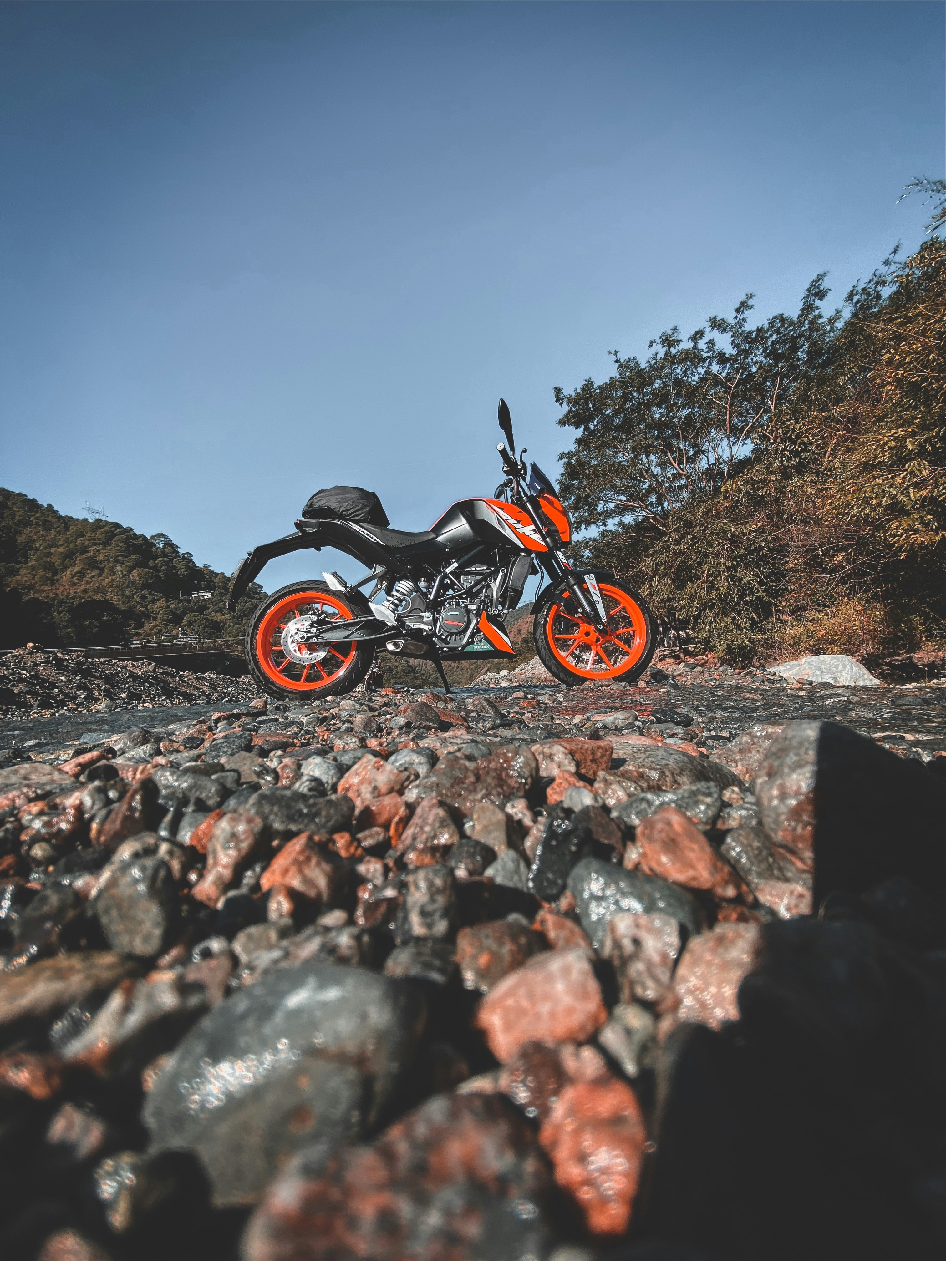 Orange-Rimmed Moto on a Rock-Strewn Riverbank Under a Clear Sky