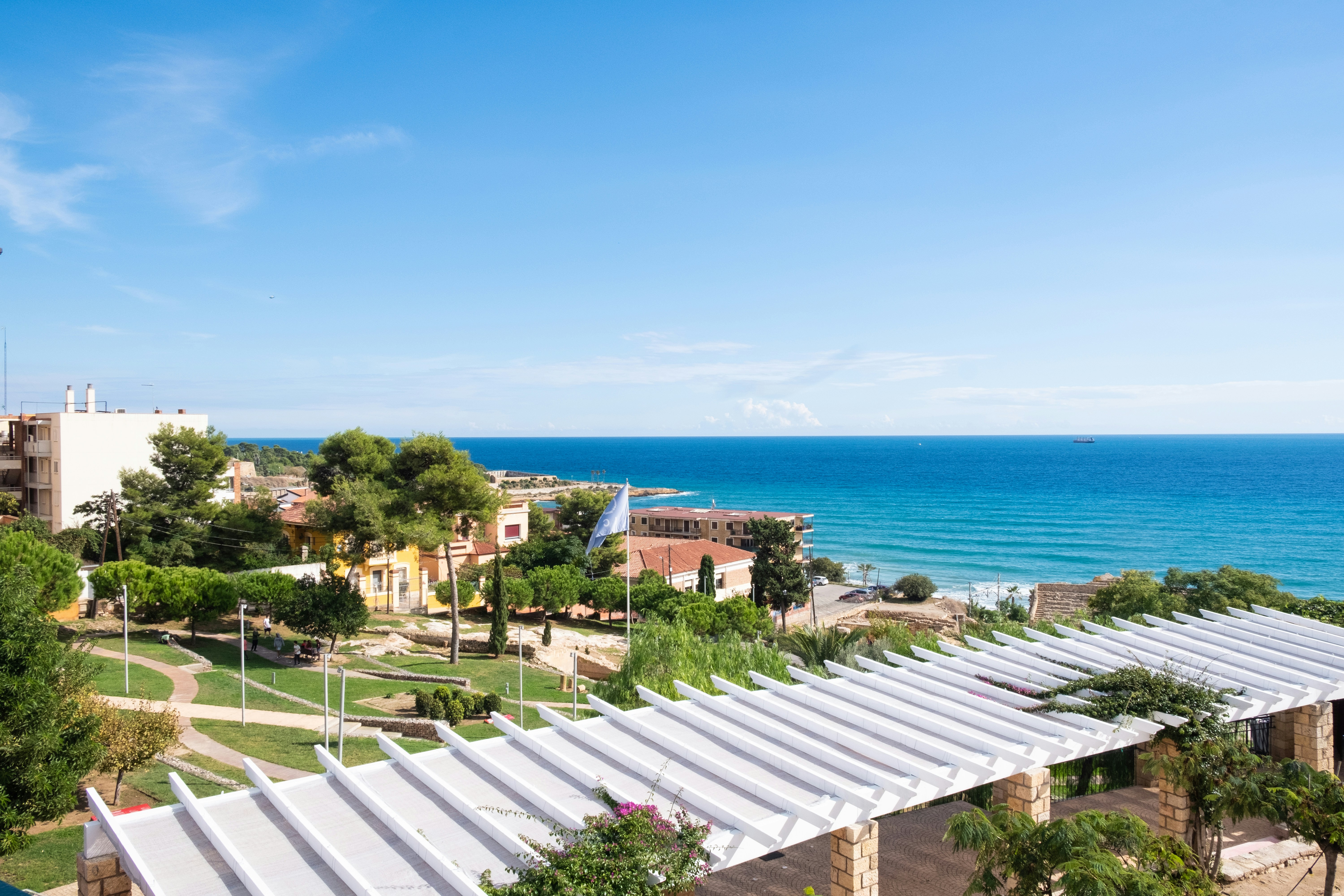 Coastal landscape with clear blue skies and turquoise sea, framed by lush greenery and modern architecture.