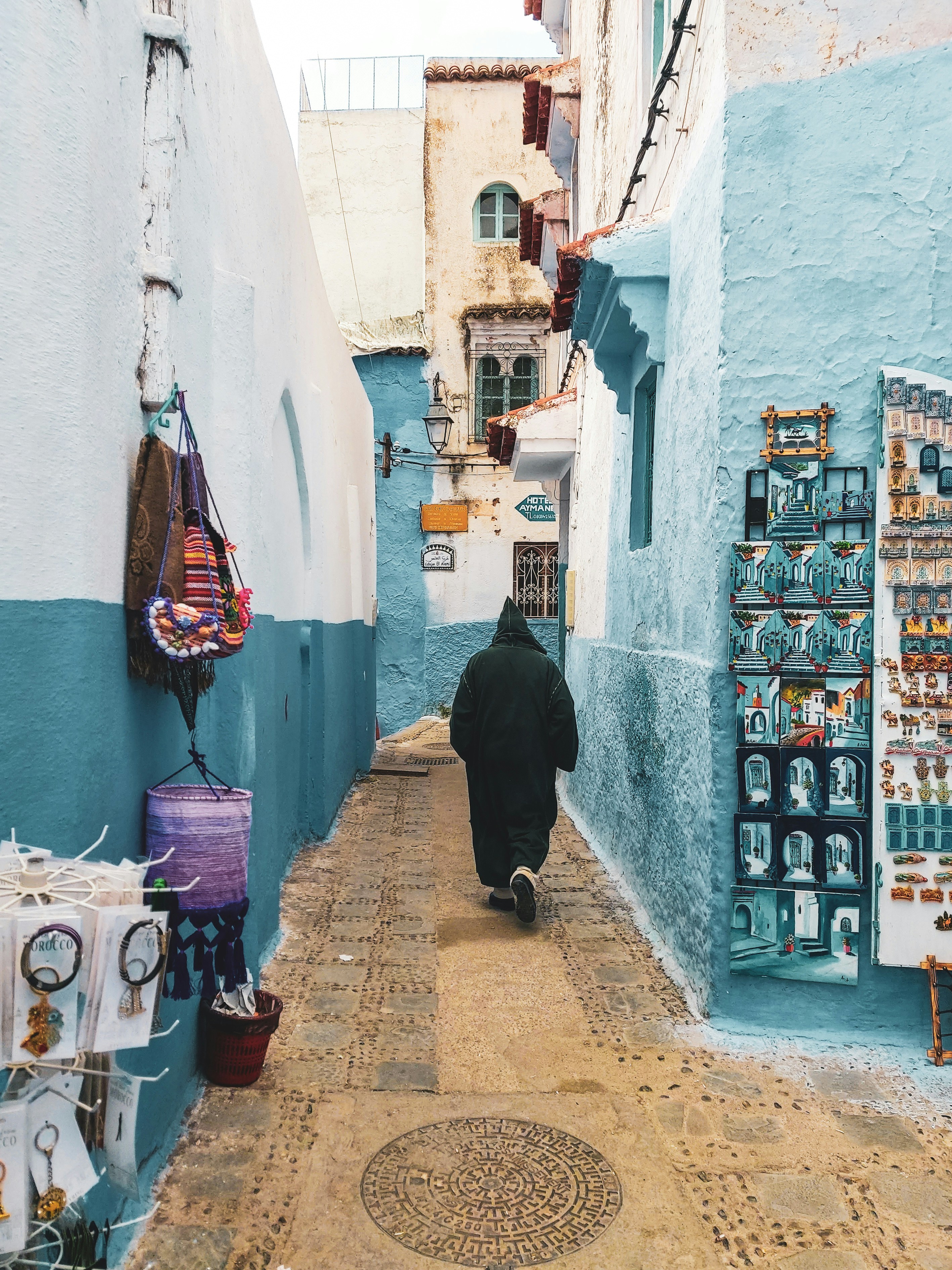 A solitary figure in a dark cloak walks through a narrow, vibrant blue alley, flanked by colorful artisan crafts and intricate wall textures.