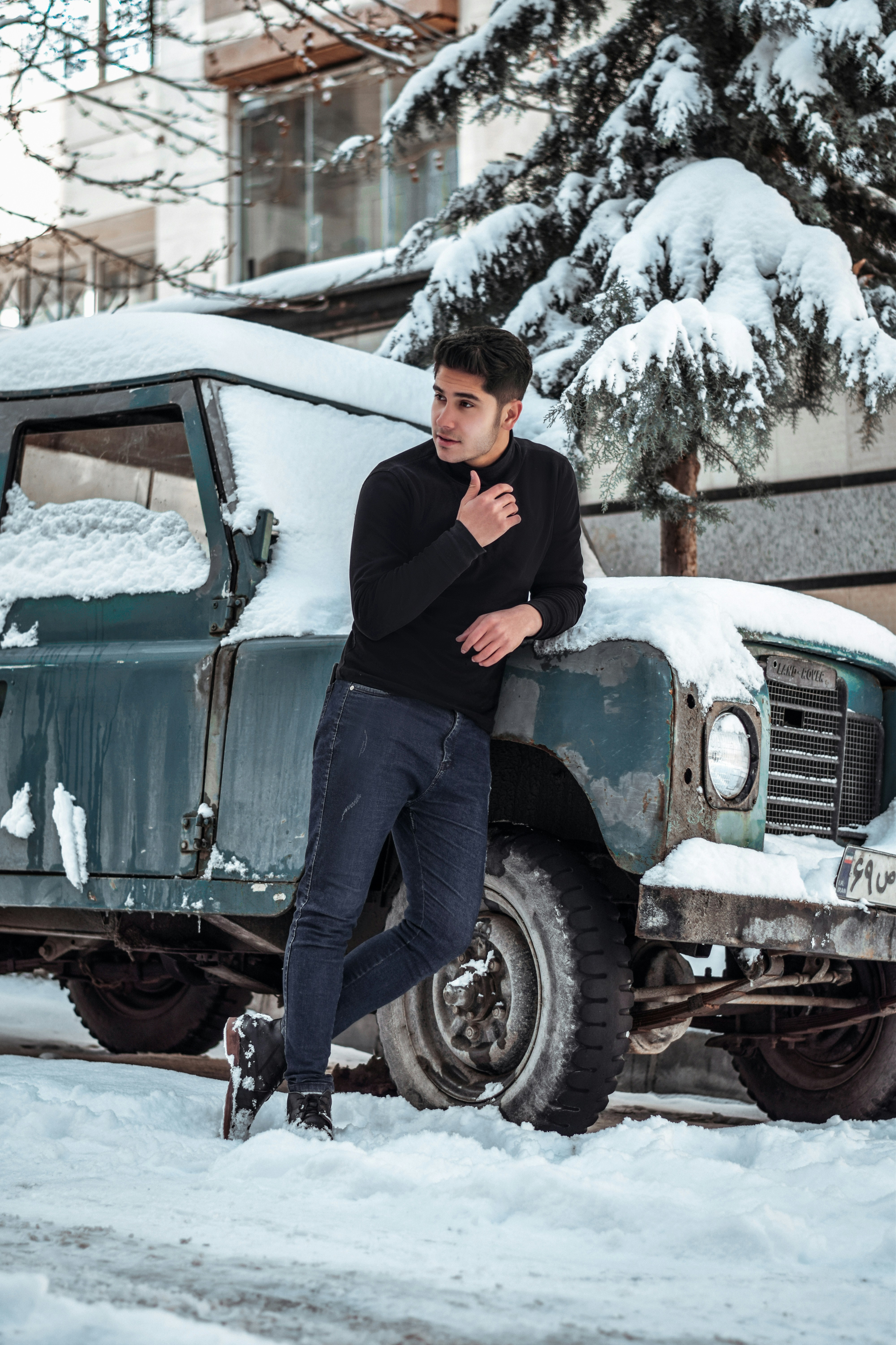 Young man leans against a snow-covered vintage truck, dressed in dark sweater, as winter light softens the scene. The image emphasizes mood and urban isolation with a hint of nostalgia.