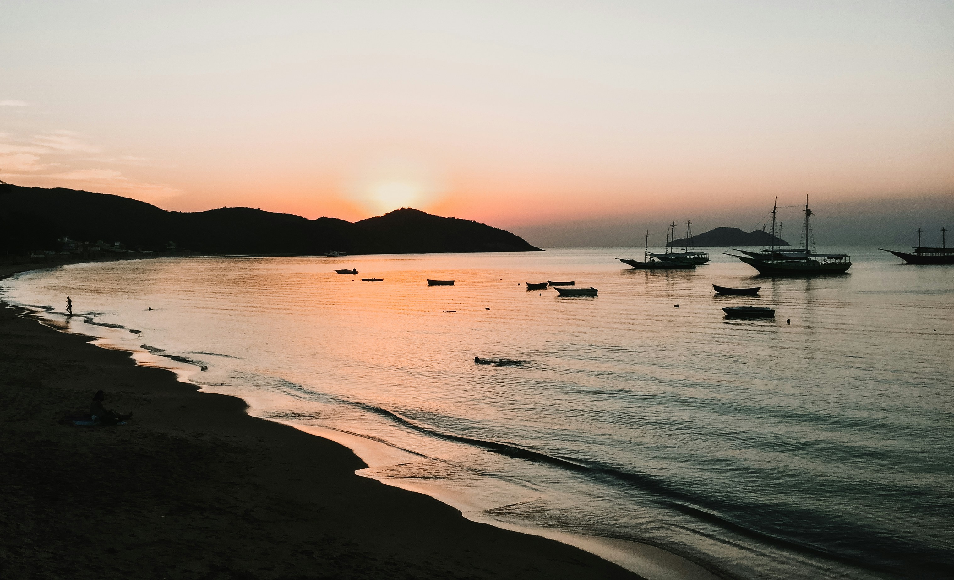 Silhouetted boats anchored in calm waters against a mountainous sunset backdrop.