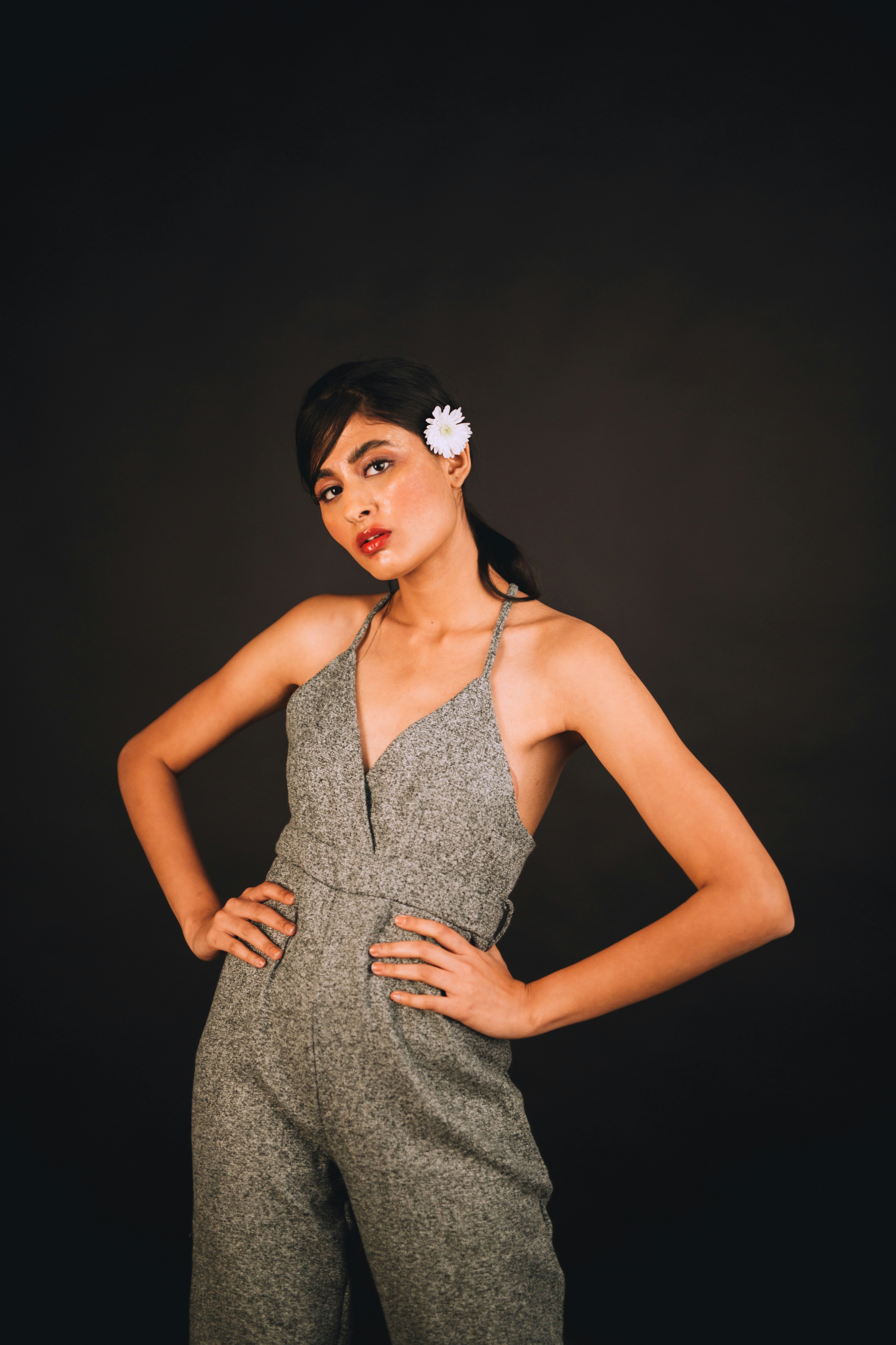 Model posing confidently in a stylish gray jumpsuit against a dark backdrop, accented by a delicate flower in her hair.