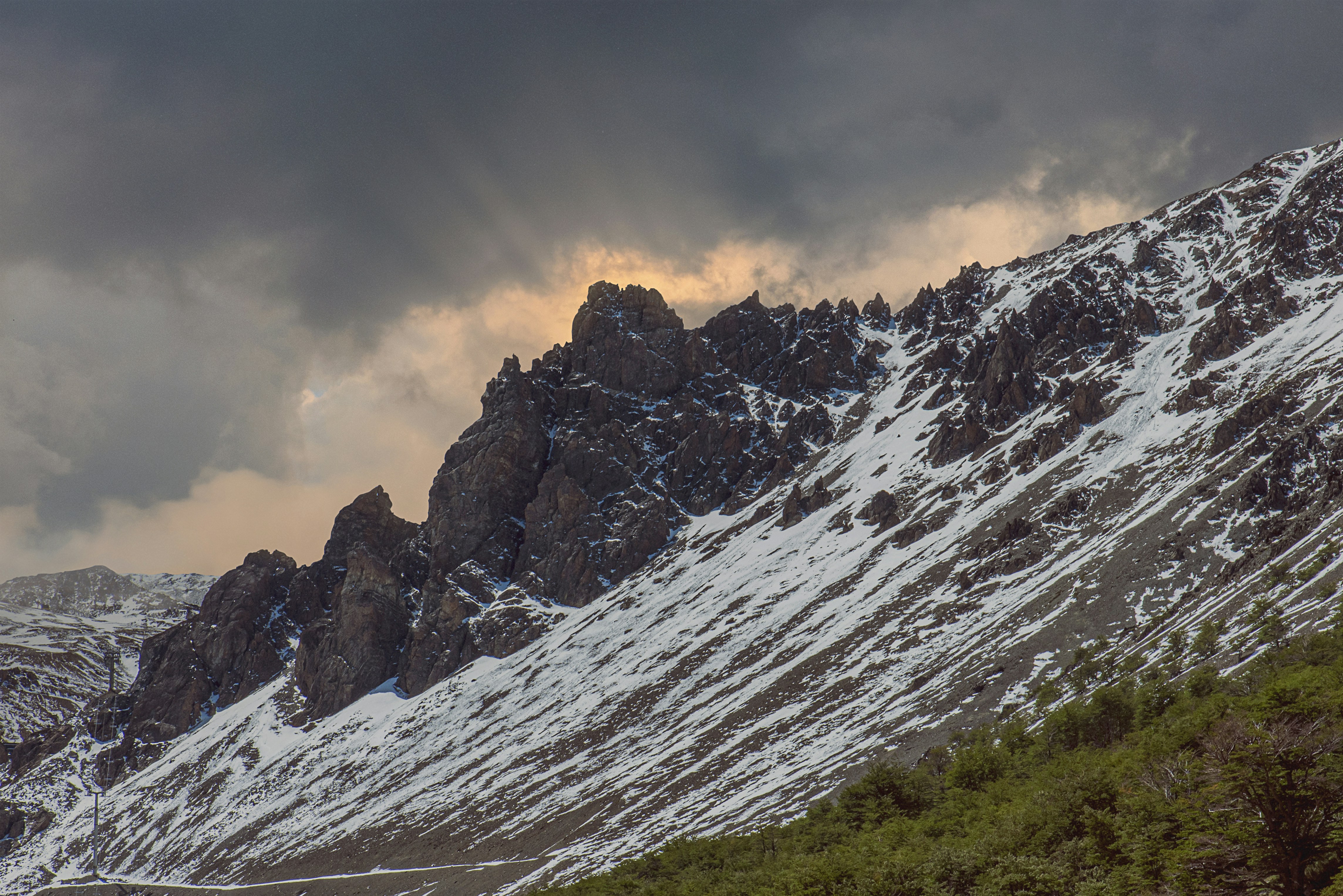 Dramatic mountain range with jagged peaks partially covered in snow, illuminated by rays of light breaking through dark clouds.