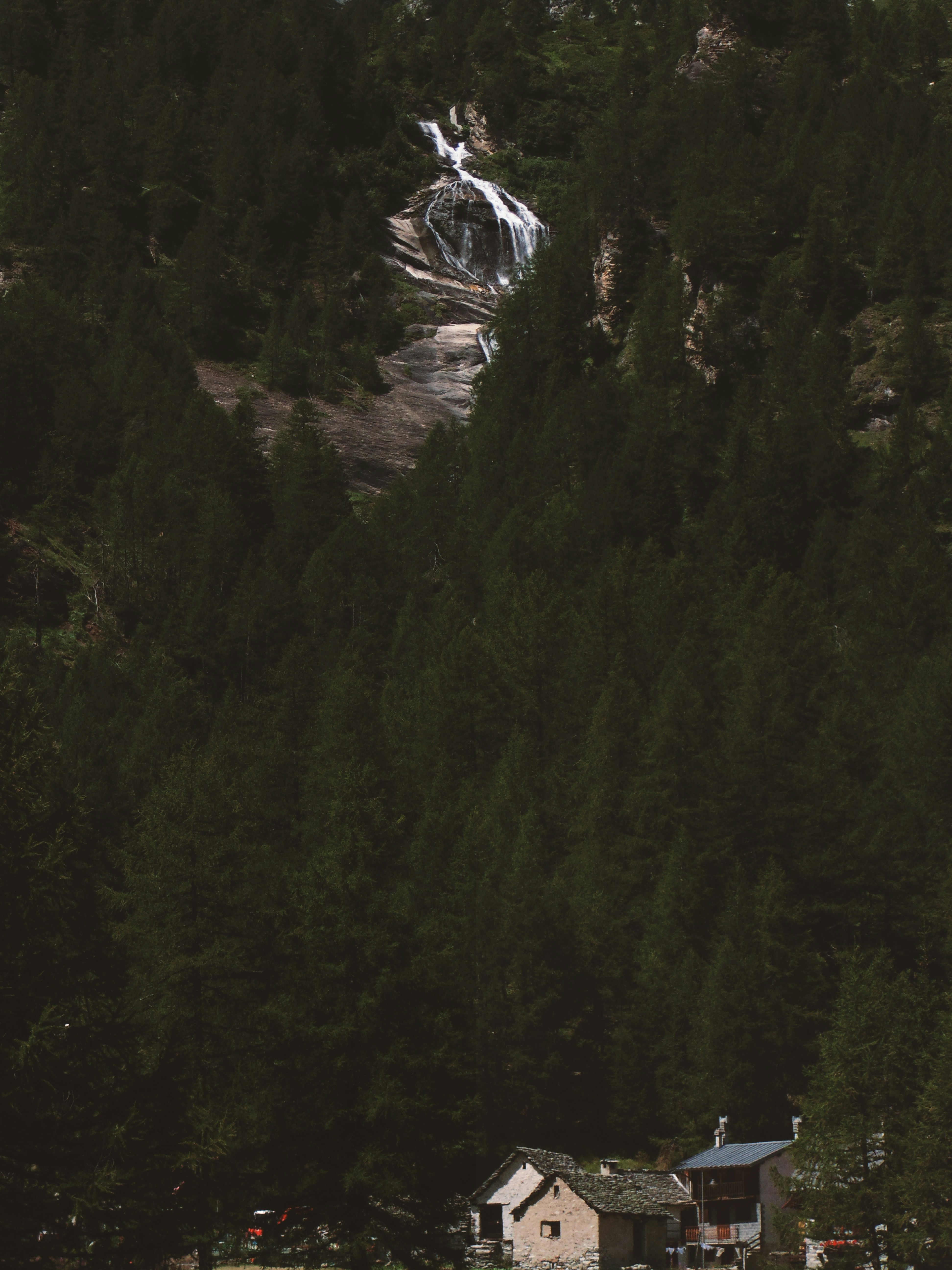 Waterfall cascading down a rocky cliff surrounded by dense forest, with a small house nestled in the foreground.