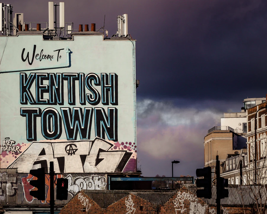 Large mural reading “Welcome to Kentish Town” on a pale building, with rooftops and industrial antennas below. Dark dramatic clouds fill the sky to the right.
