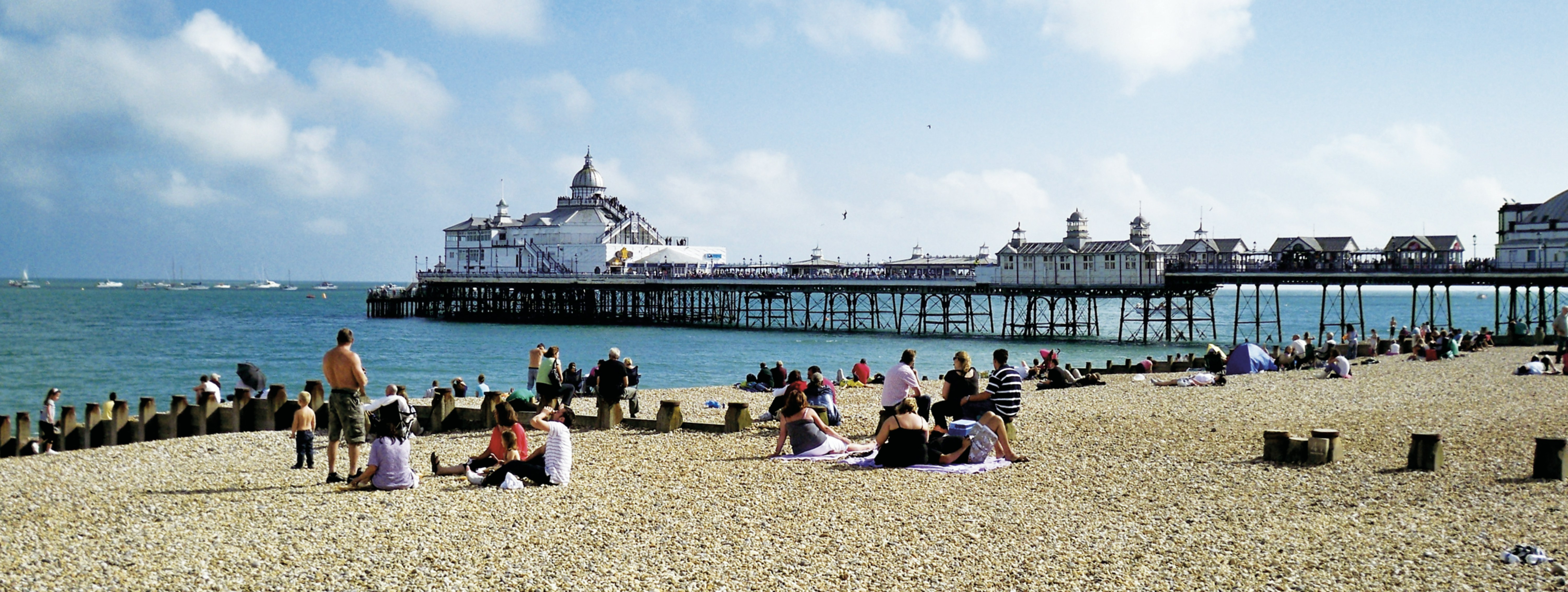 People relaxing on a sandy beach with Eastbourne Pier extending into the sea under a partly cloudy sky.