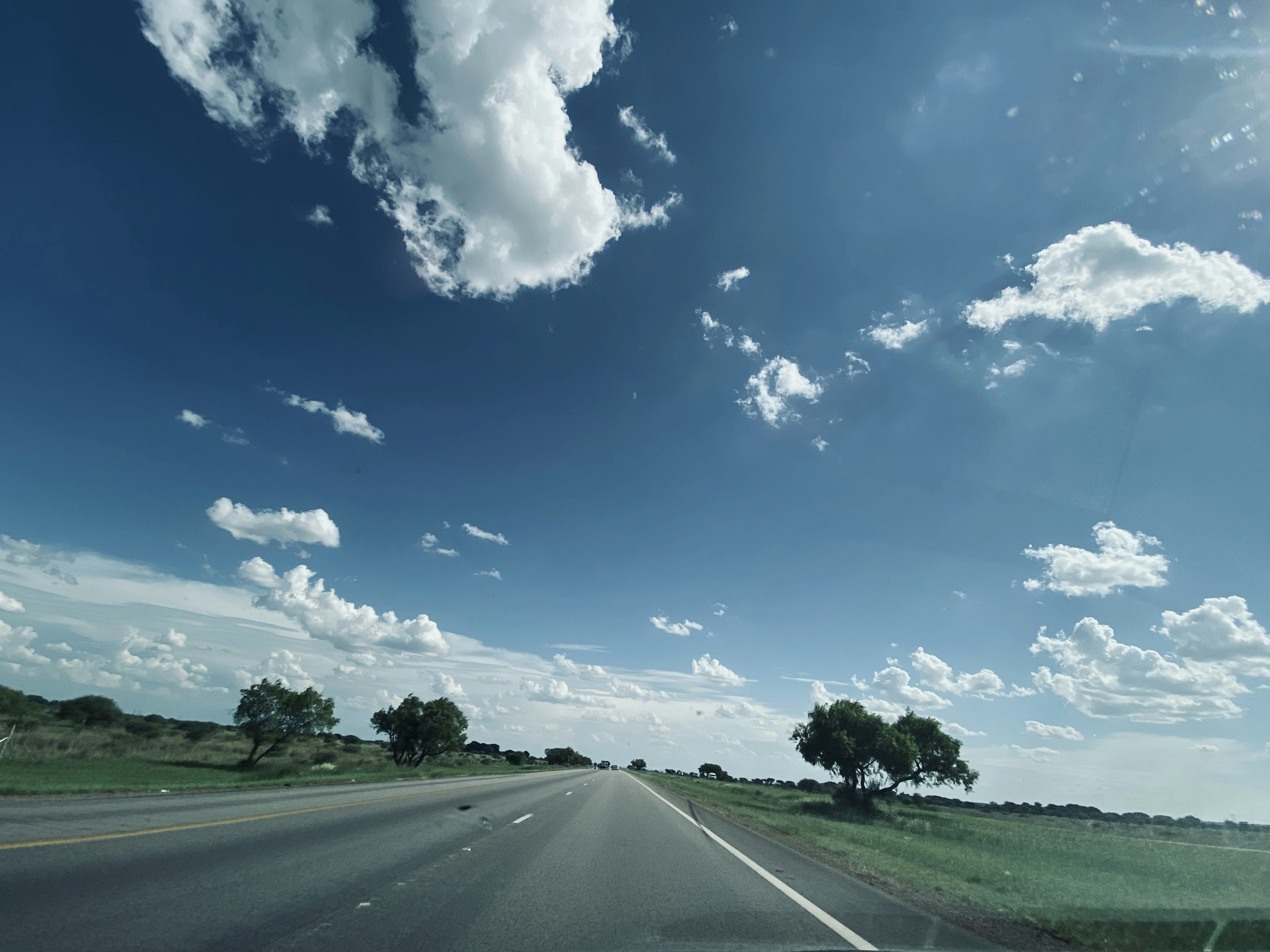 Wide-open highway stretching into the distance, flanked by lush greenery and scattered trees under a vibrant sky filled with clouds.