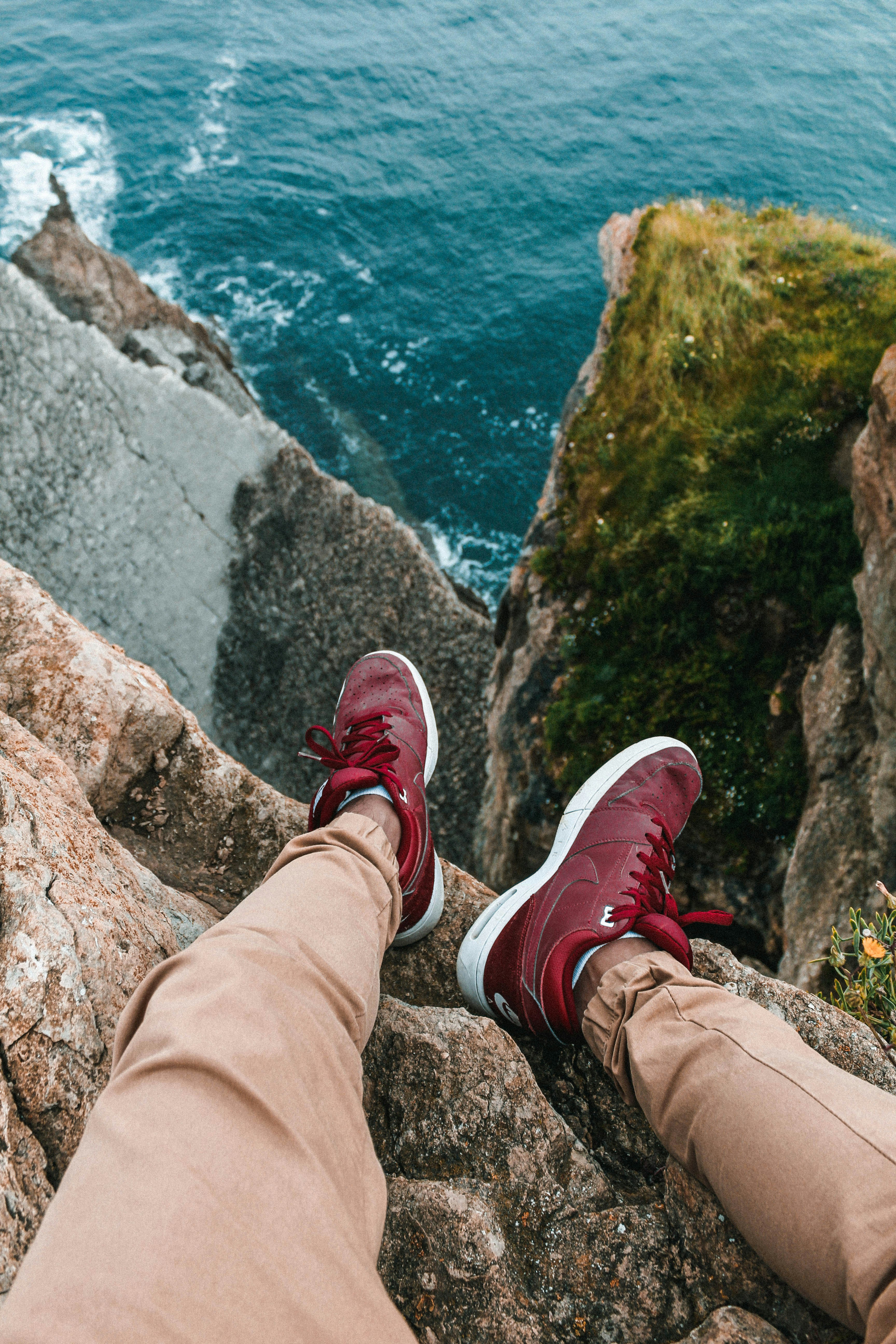 Person sitting on rocky cliff edge overlooking blue ocean below.