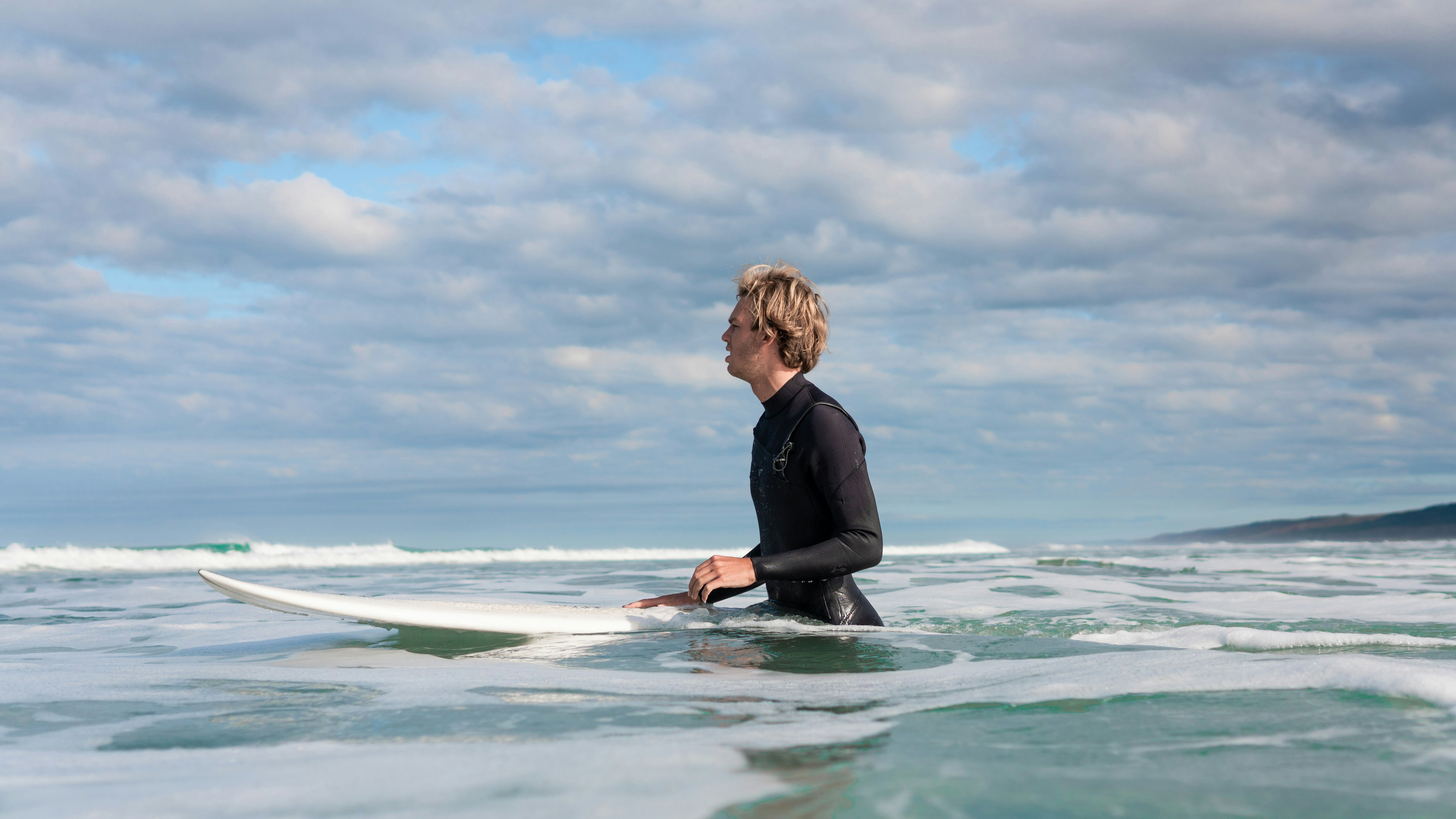 Surfer poised in the ocean, holding a surfboard amidst gentle waves under a cloudy sky.