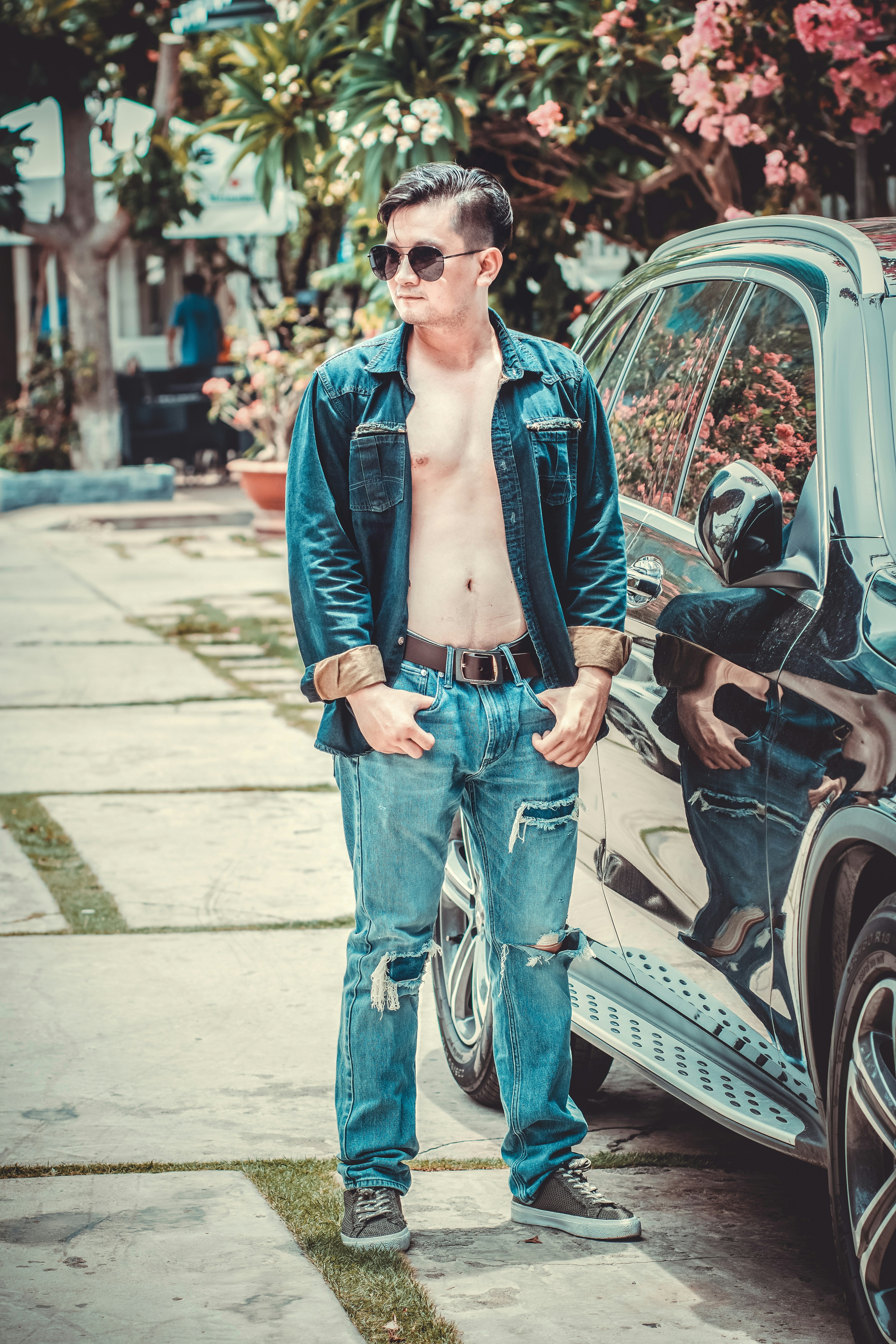 A young man in a denim jacket and ripped jeans poses confidently beside a sleek car, surrounded by vibrant floral greenery.